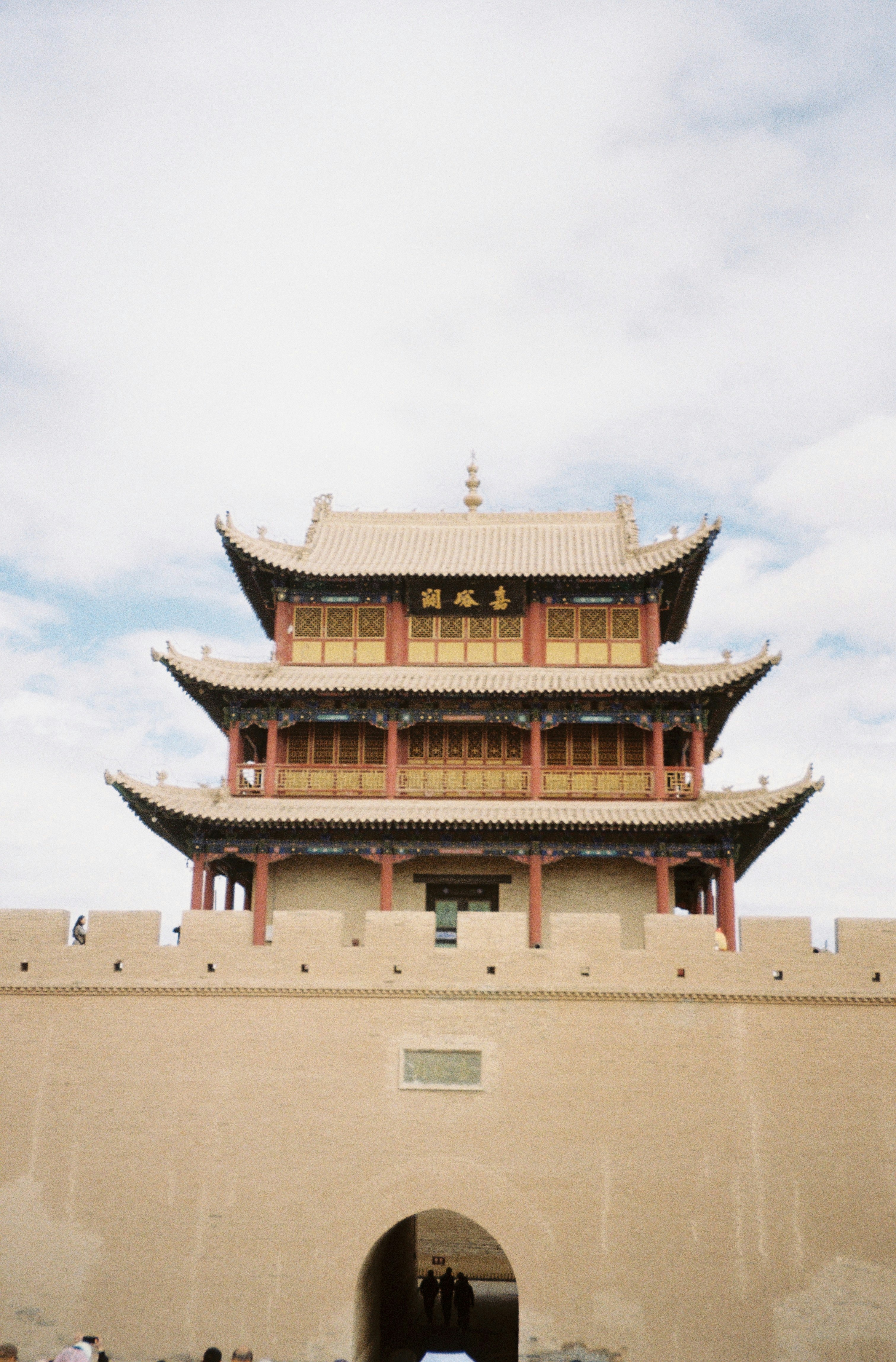 Three-tiered pagoda atop a fortified gate rises above a sunlit brick wall, set against a bright blue sky; a gateway tunnel anchors the foreground for a sense of scale.
