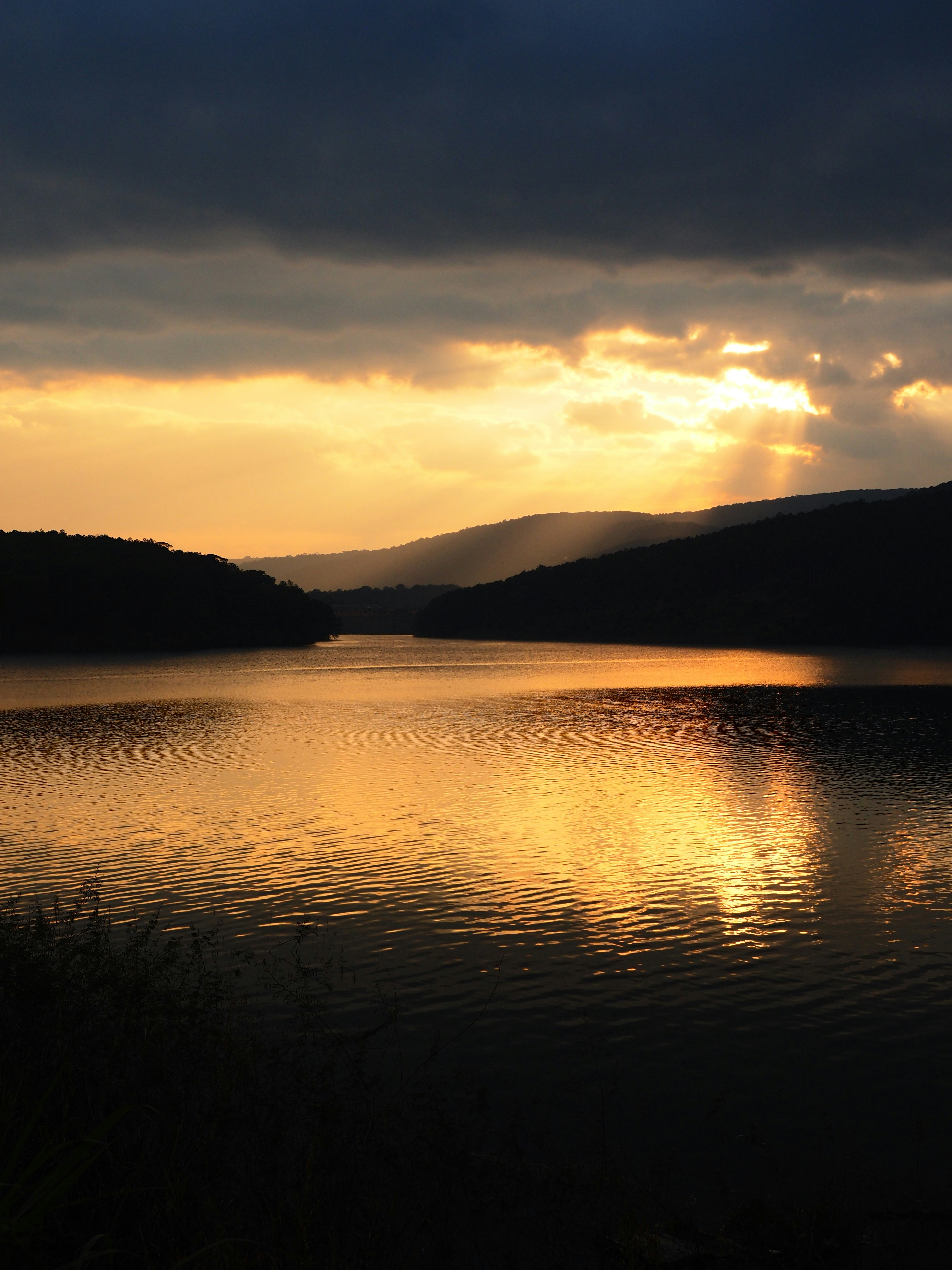 Warm sunset over a calm reservoir with dark silhouettes of hills; sunlight breaks through clouds, lighting the water with a silvery-gold sheen. A tranquil landscape shot capturing the moment before twilight; note: photographic scene, not a logo or illustration.