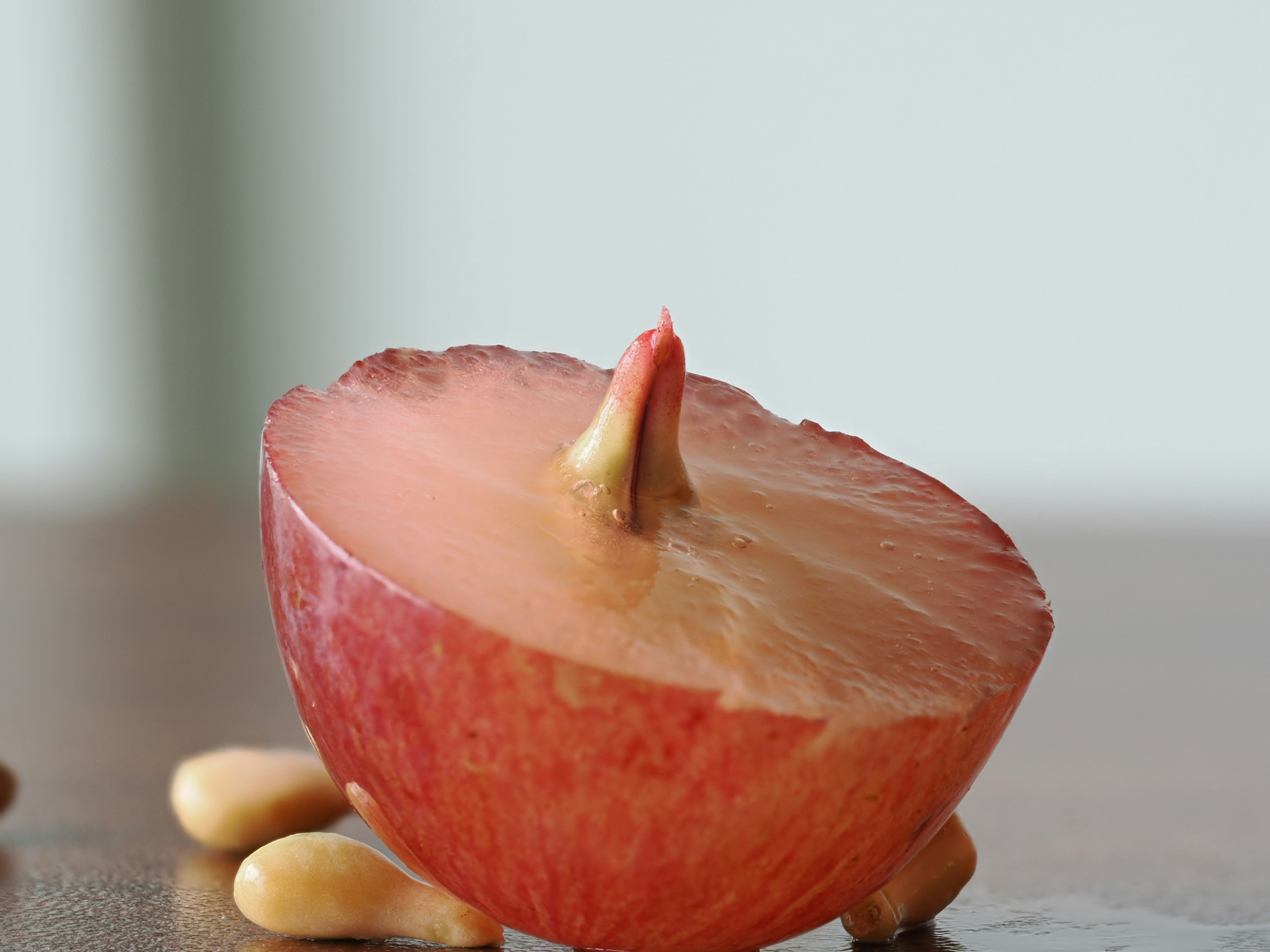 Halved red apple with exposed flesh resting on a wooden table, some pale seeds nearby, soft background blur.
