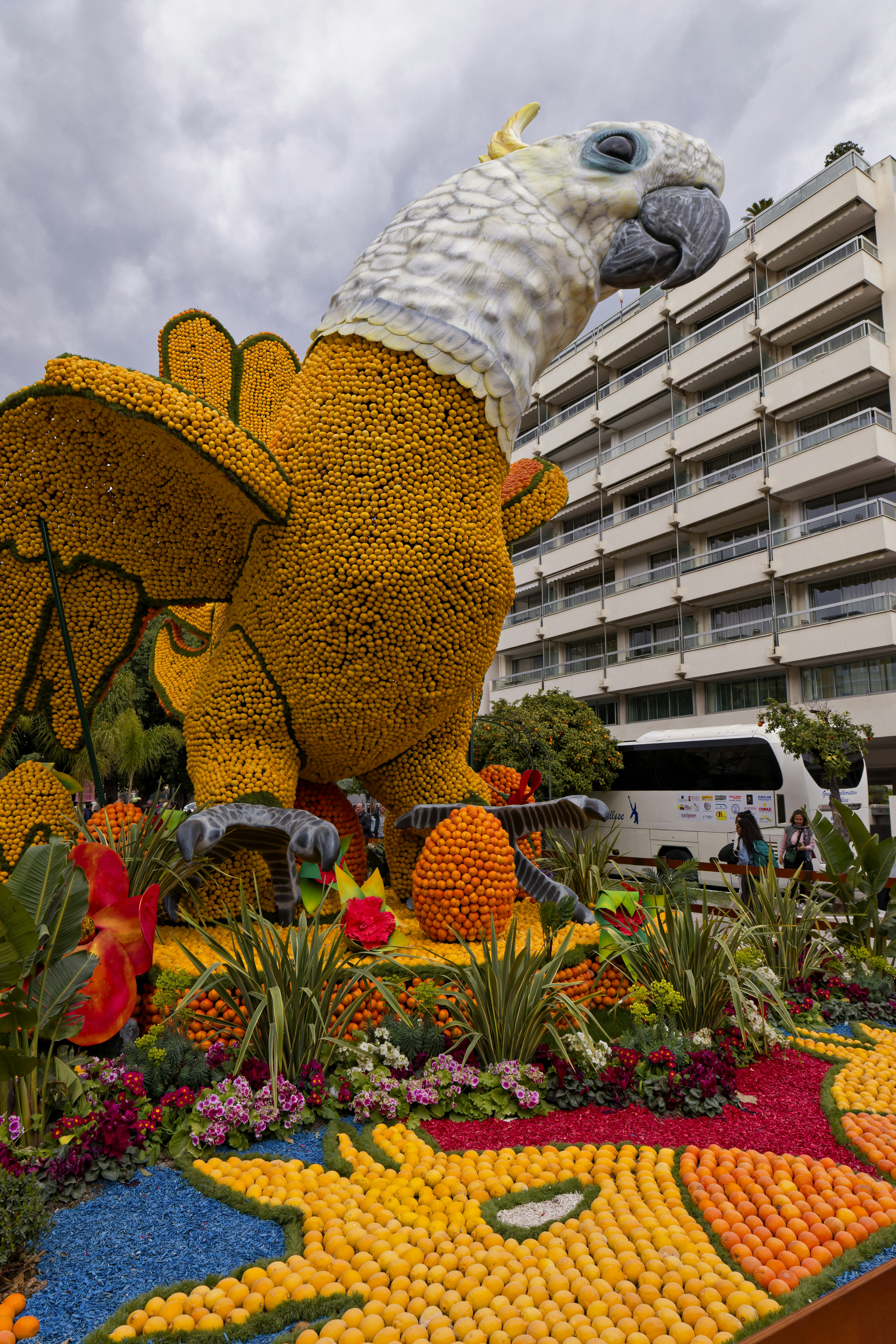 A colossal dragon sculpture made of orange flowers sits in a city garden, with a modern apartment building in the background and bright flower beds in the foreground.