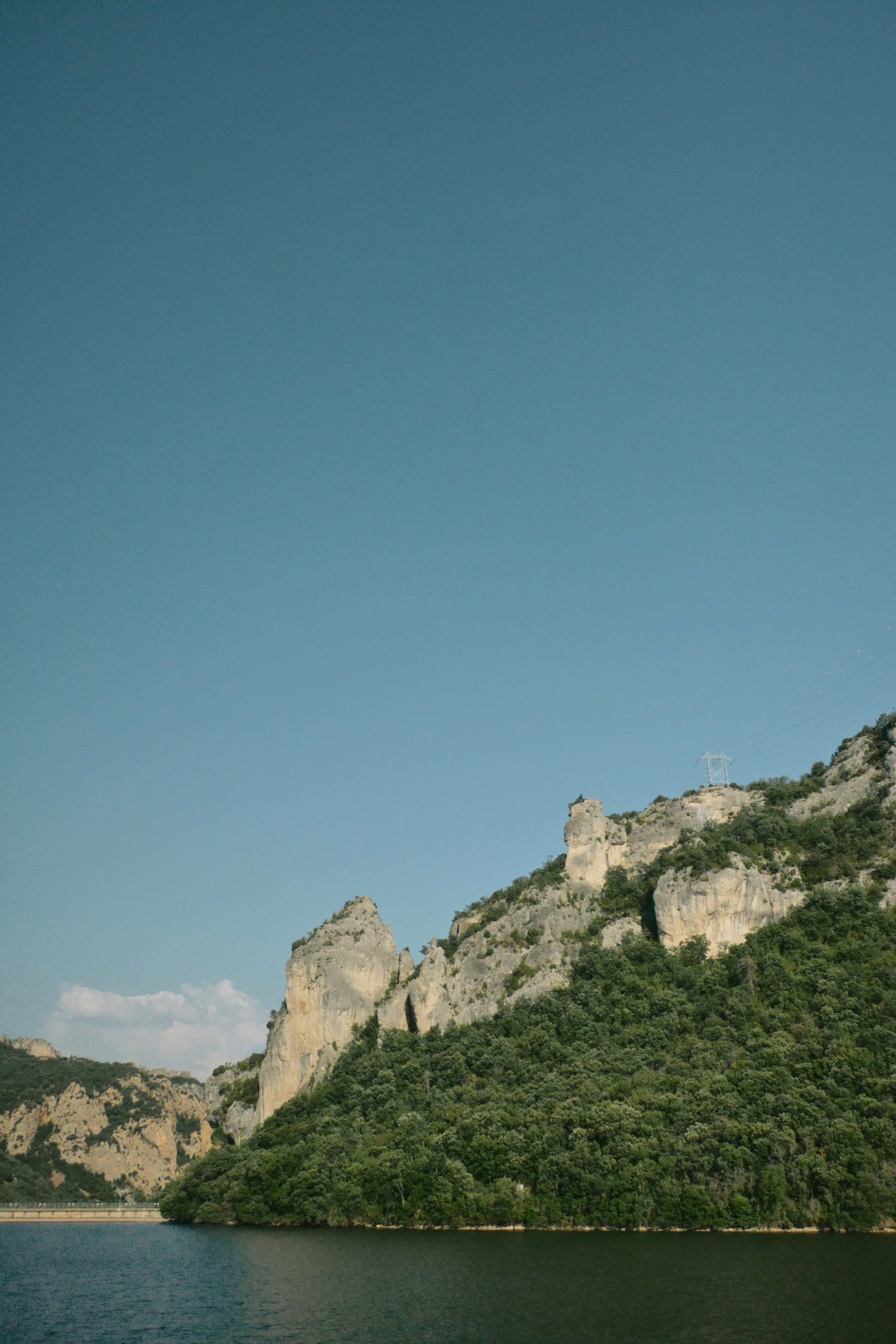 Sunlit rocky cliffside and treeline along a calm coastal inlet under a blue sky. A natural landscape scene without people, showcasing rugged geology and tranquil water. This is a photographic capture of a serene coastal outcrop.