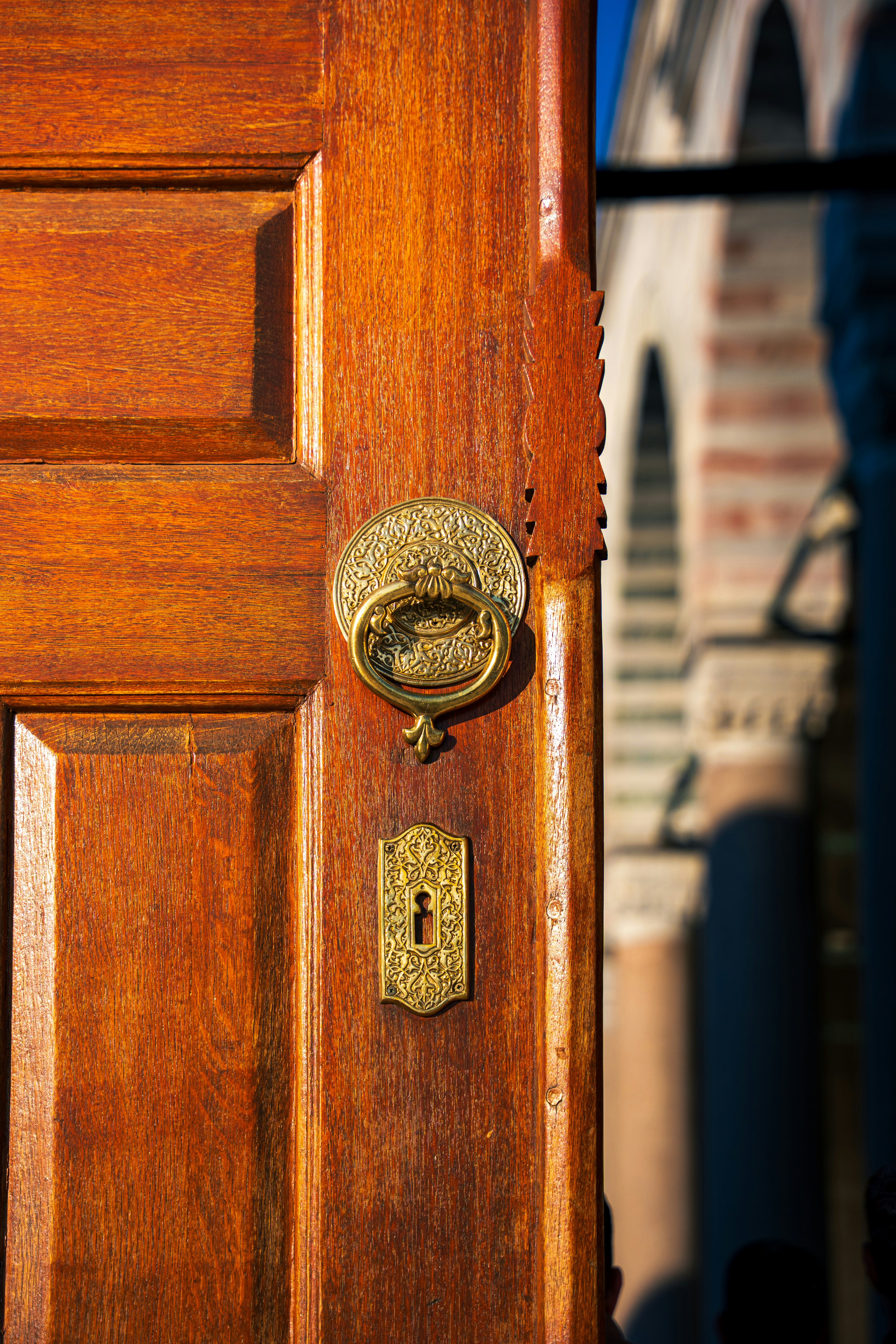 Close-up of an ornate brass door knob and keyhole mounted on a rich wooden door, with architectural detail and brickwork faintly visible in the background. The scene emphasizes texture, craftsmanship, and a sense of entry.
