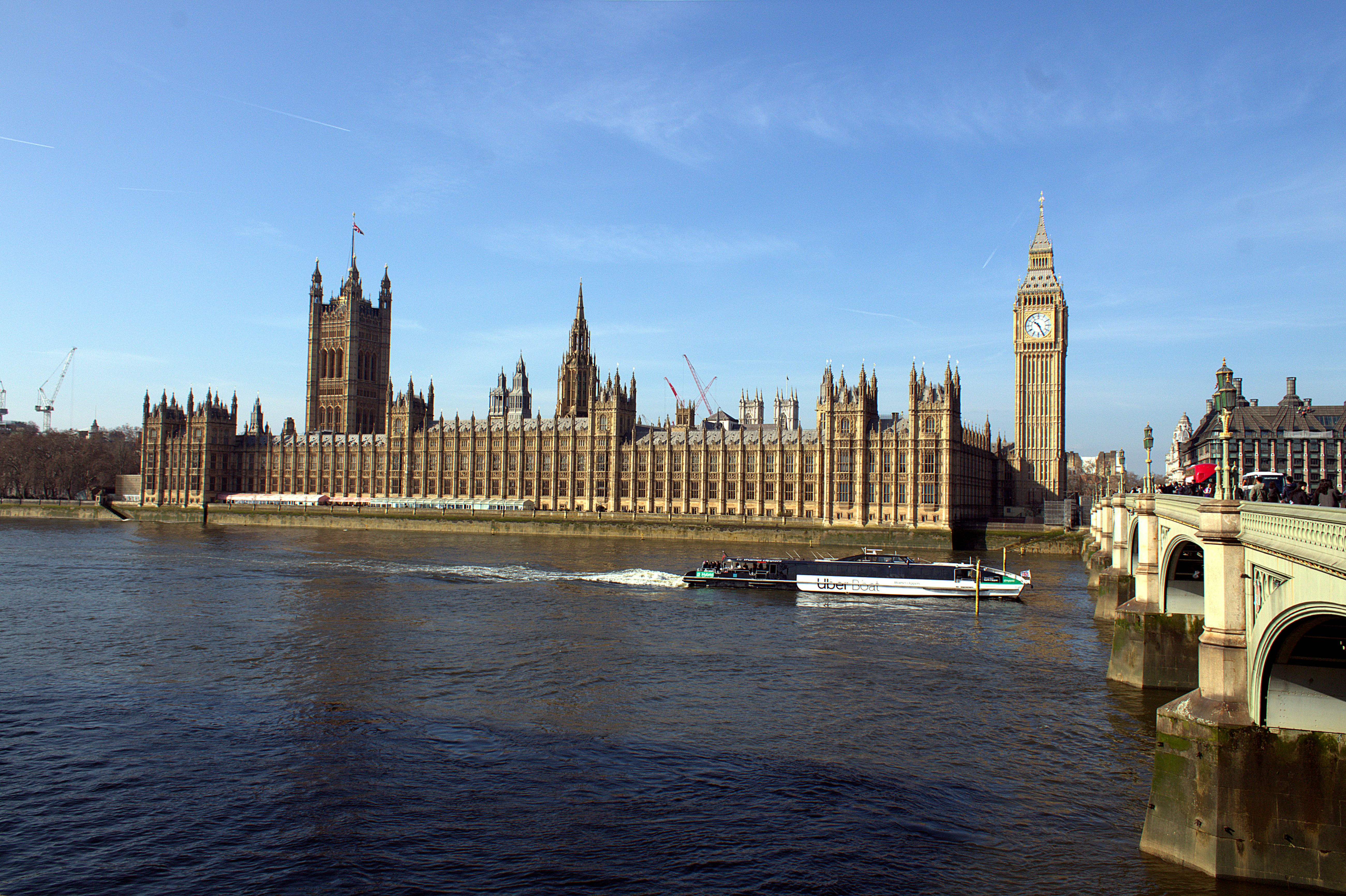 Wide view of the Thames with the Houses of Parliament and Big Ben in daylight; a boat glides along the river near Westminster Bridge, capturing clear architectural detail and blue sky.