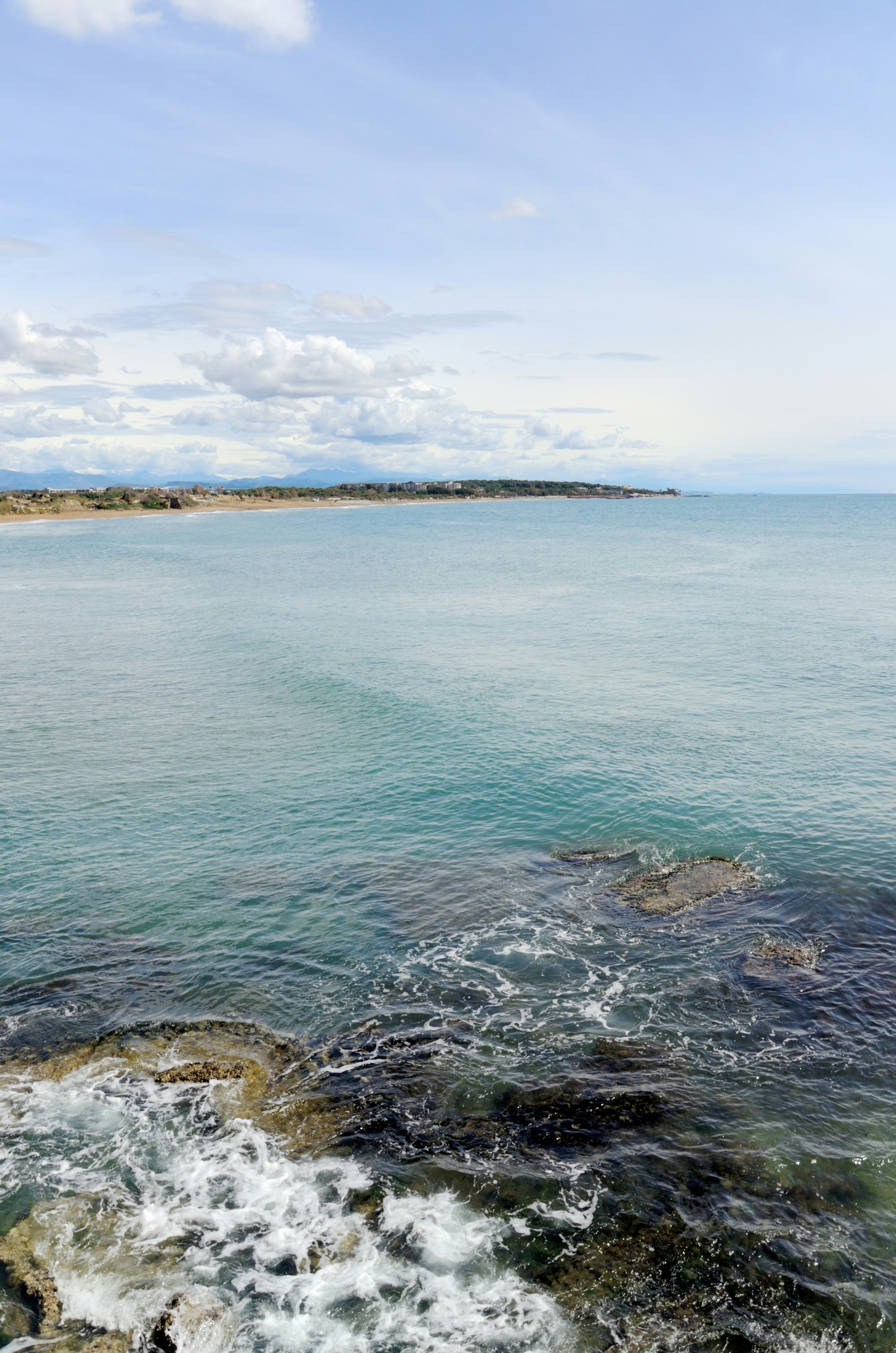 Coastal scene with clear turquoise water lapping over rocks, a distant shoreline under a blue-sky horizon. A calm seascape, highlighting the translucent surf and shallow rocky seabed.