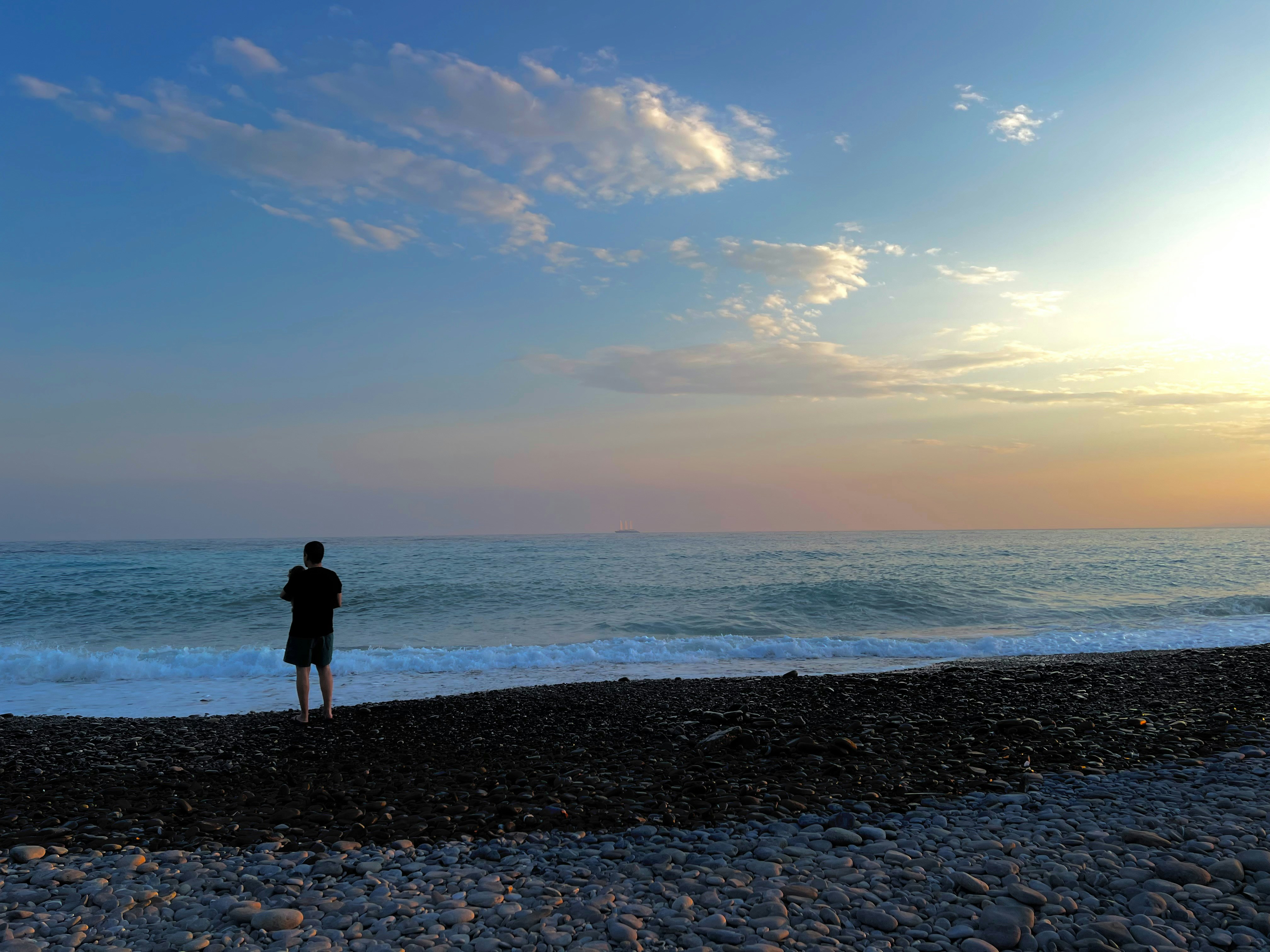 A lone person stands at the pebble beach facing the sea during sunset, with a calm horizon and soft clouds. The scene emphasizes quiet solitude and coastal texture; photograph is a natural landscape capture.