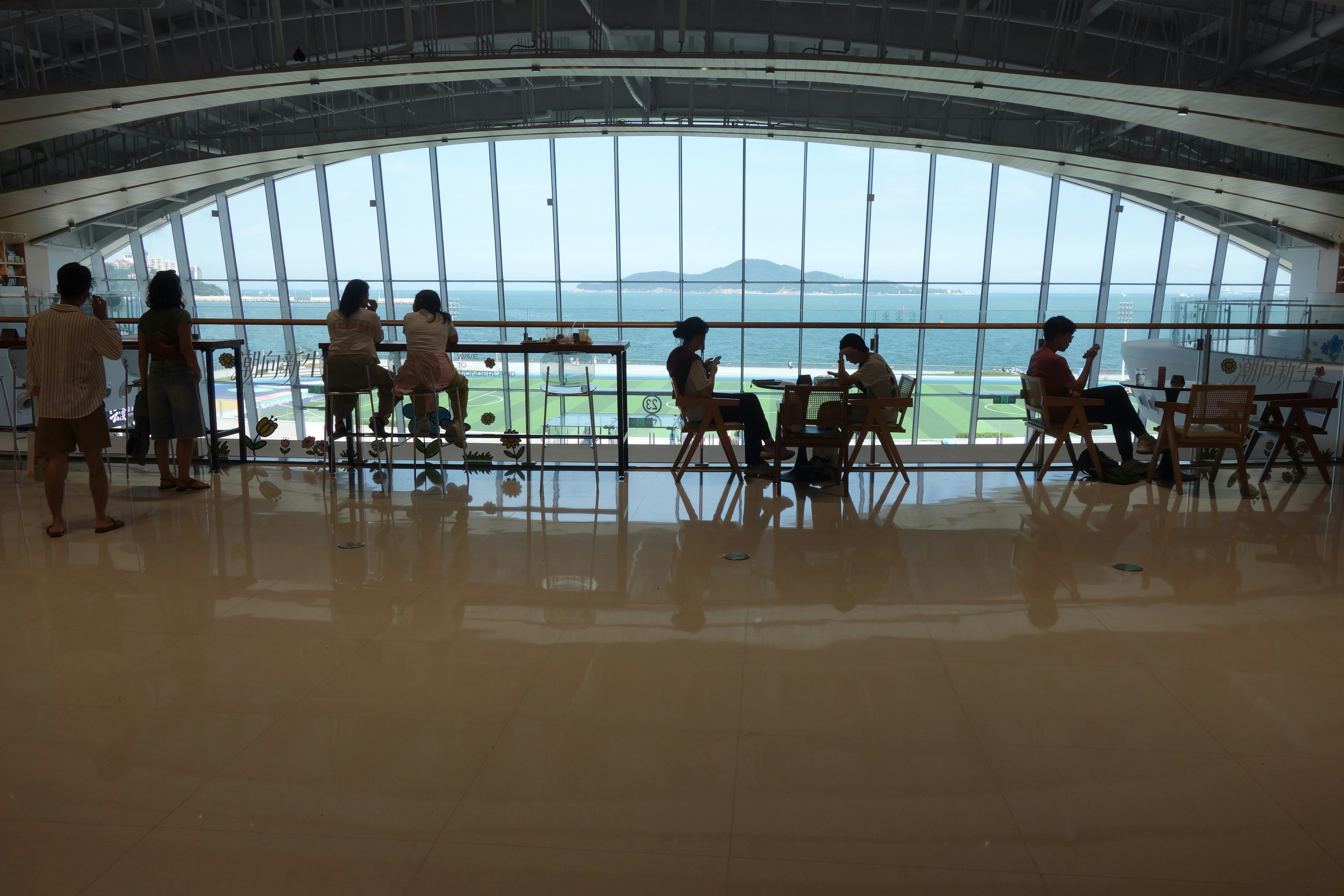Passengers at a sunlit harbor overlook inside a curved glass-terminal