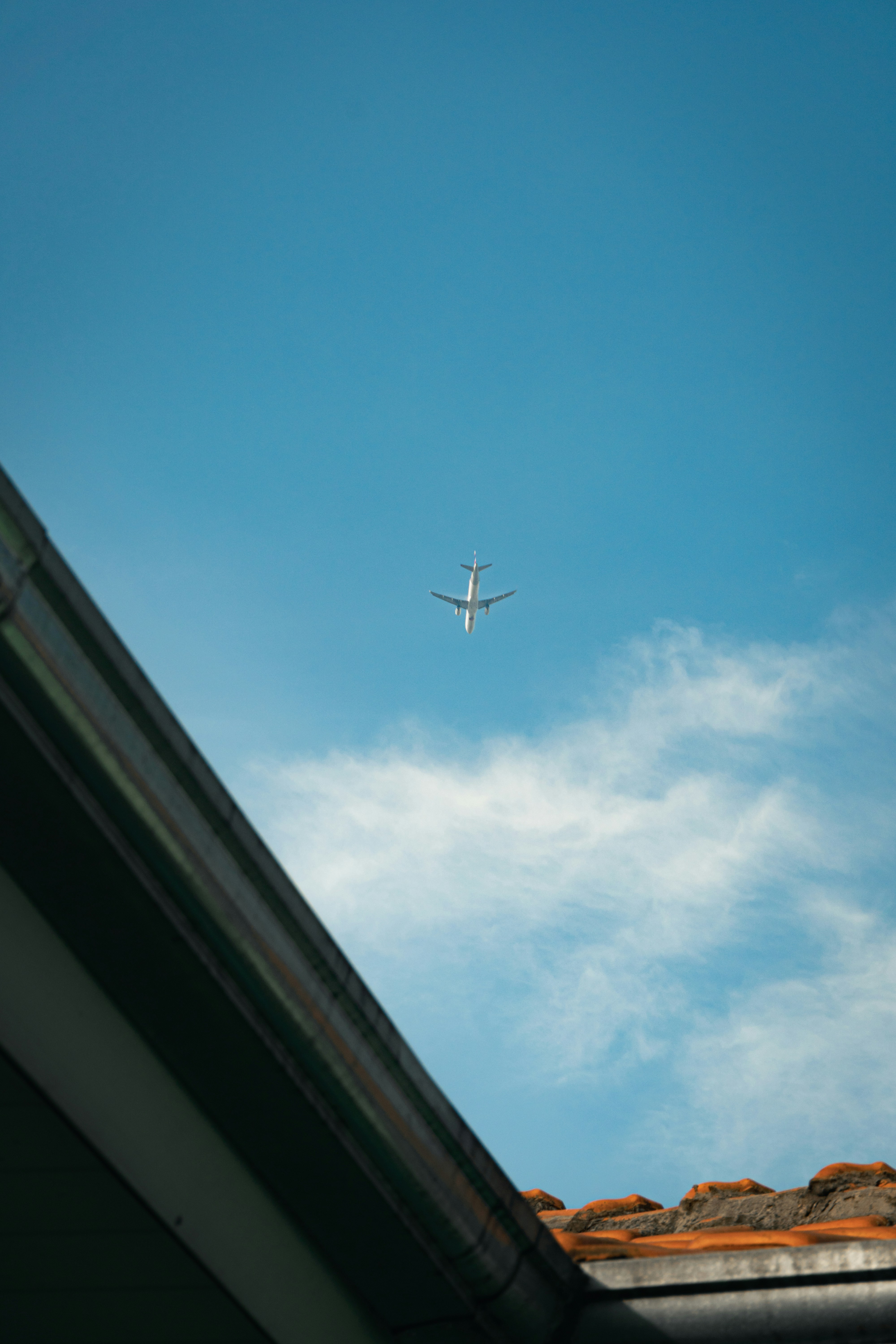 A commercial airliner sails high above rooftops against a clear blue sky, captured from a low-angle perspective. This photo focuses on the aircraft with the building edge providing a defining foreground element.