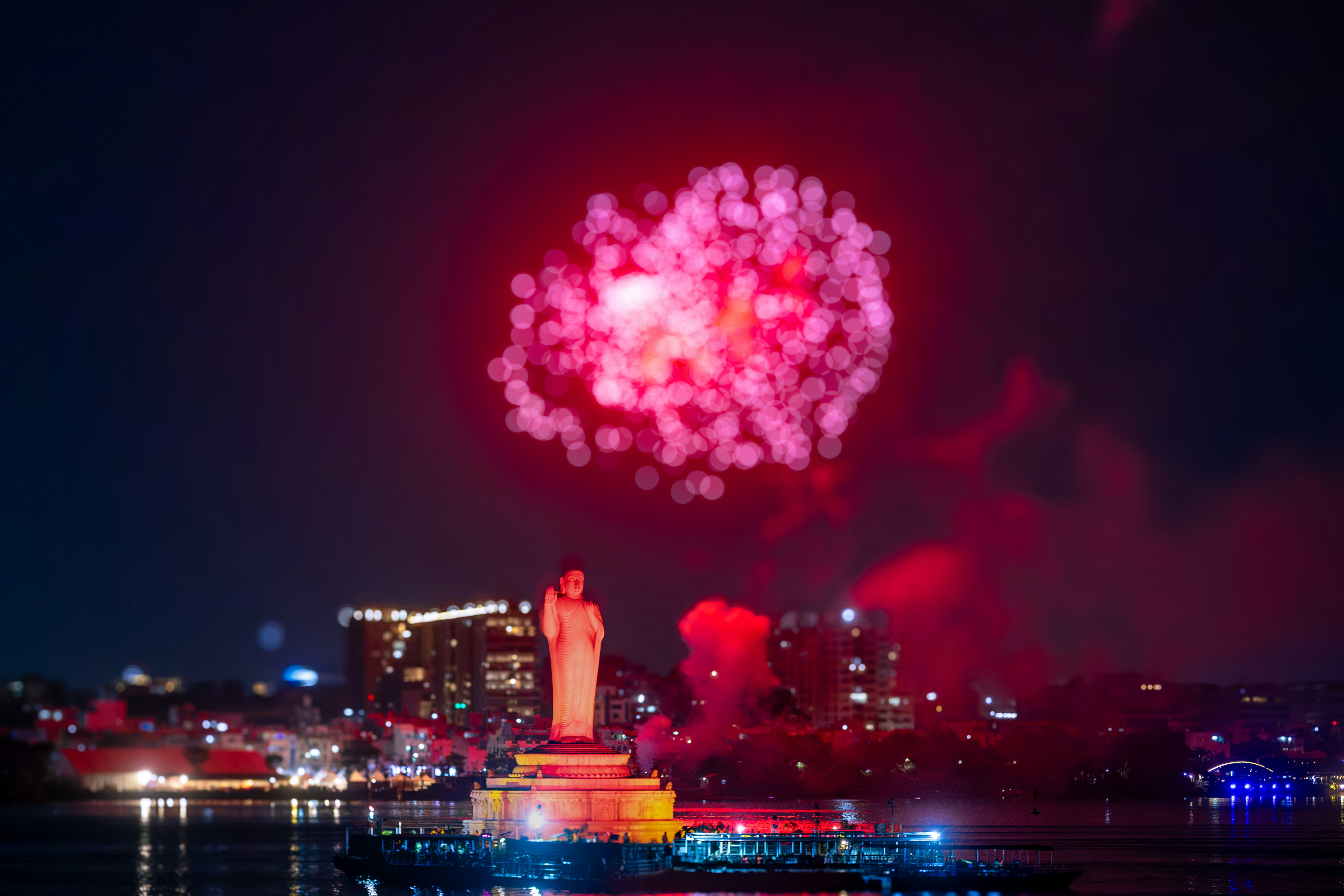 Vivid fireworks bloom above a riverside statue with city lights reflecting on the water, a nighttime urban celebration captured across a wide horizon. A commemorative or public-art scene with vibrant reds and purples.