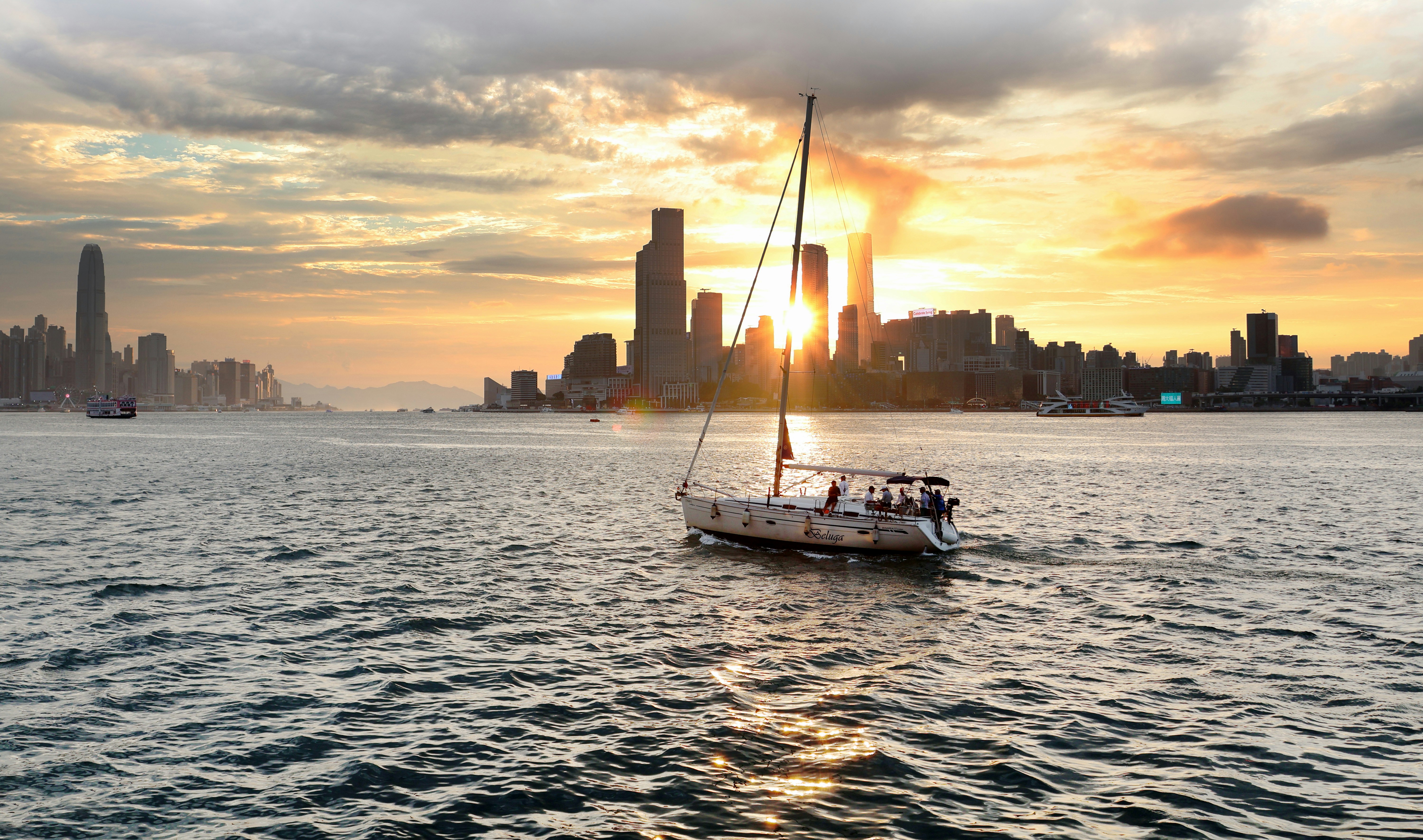 A lone sailboat glides across a calm bay with a dramatic sunset over a distant city skyline; the scene captures warm light reflecting off the water and silhouetted high-rises in the distance. (Drone-like horizon, photographic composition.)