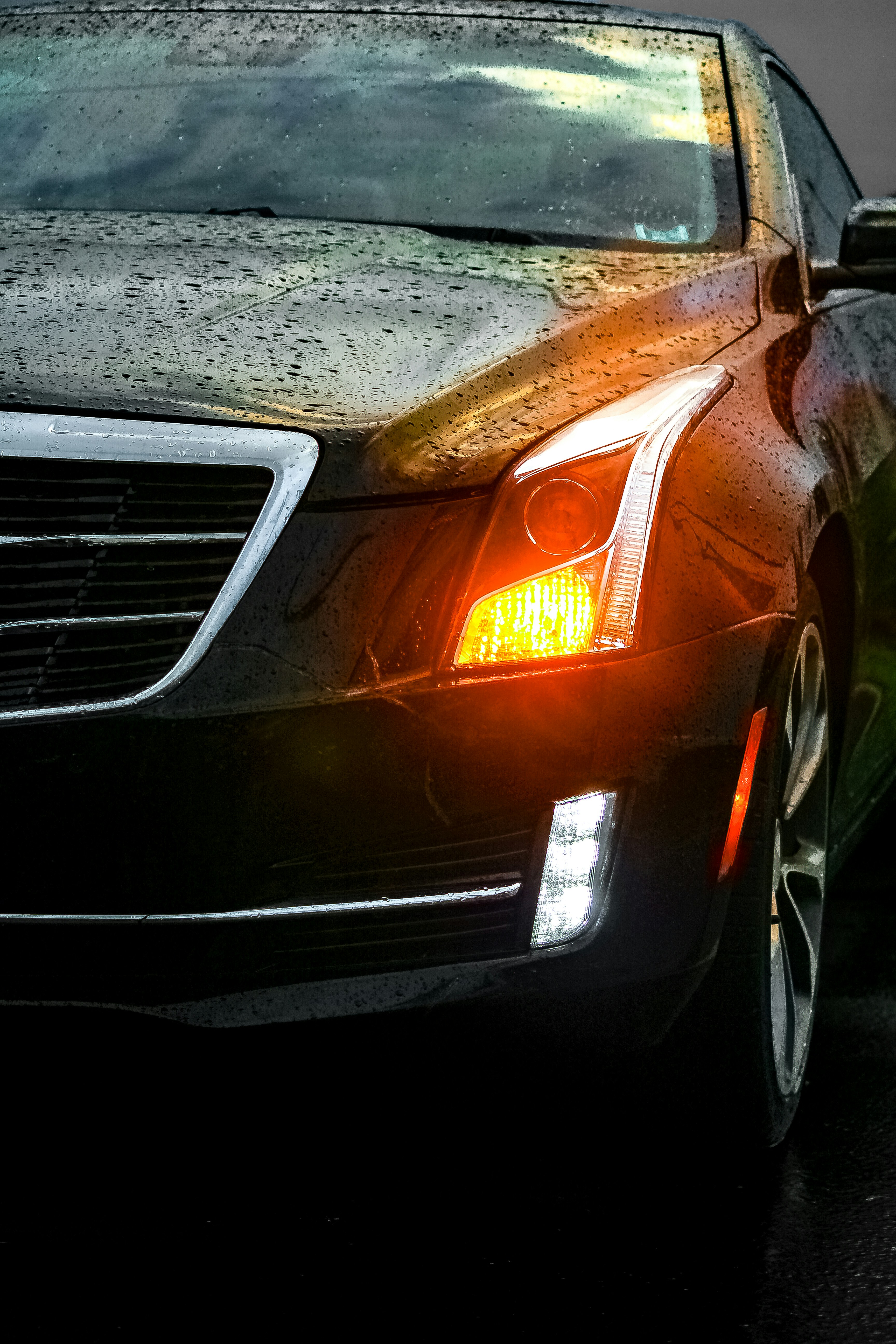 Close-up of a black luxury car front with illuminated orange turn signal and vertical daytime running light on a rain-soaked surface.