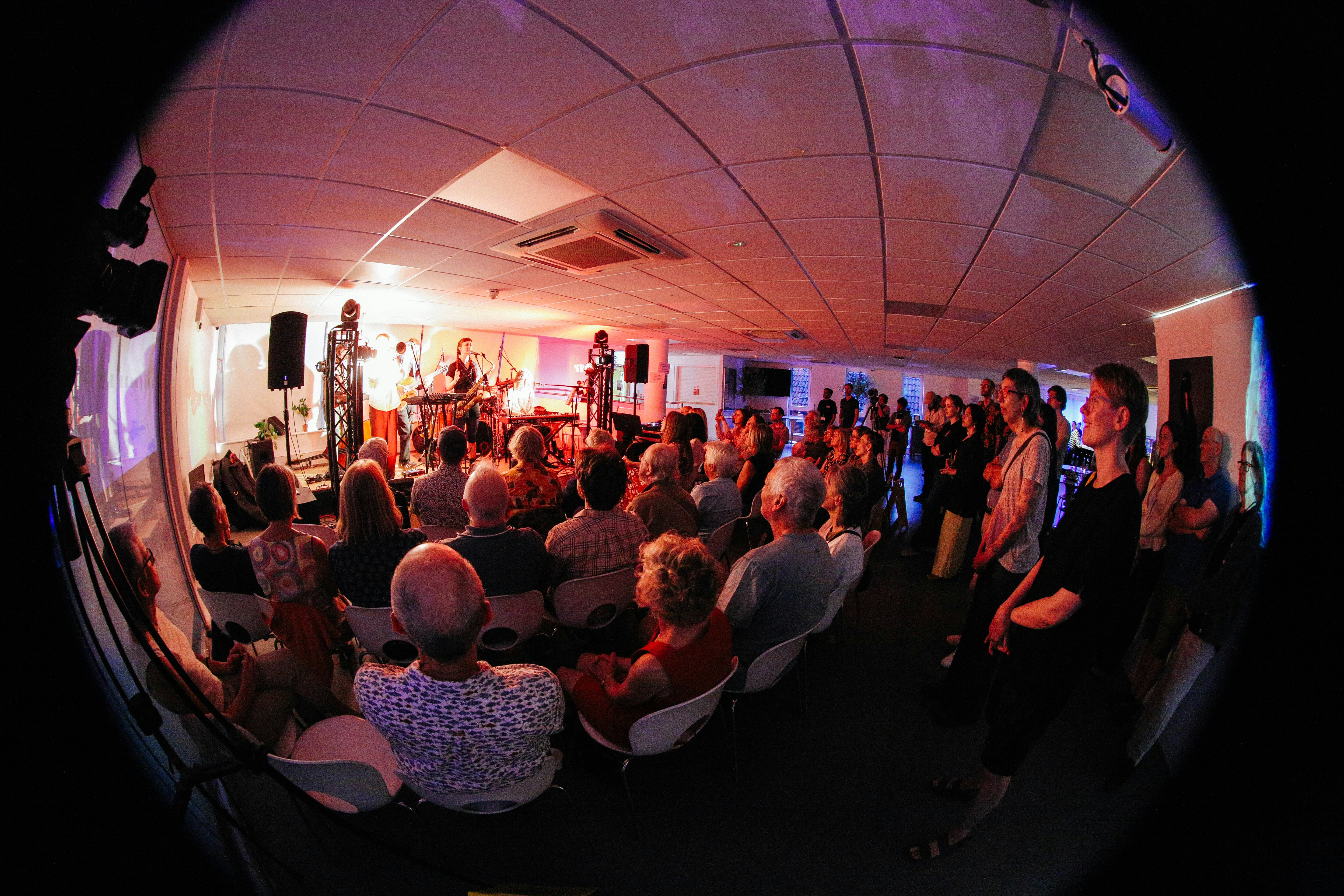 A live music performance in a compact indoor venue with an engaged audience and a bright stage; the scene includes a band, lighting, and folding chairs. This is a photo capture of the event.