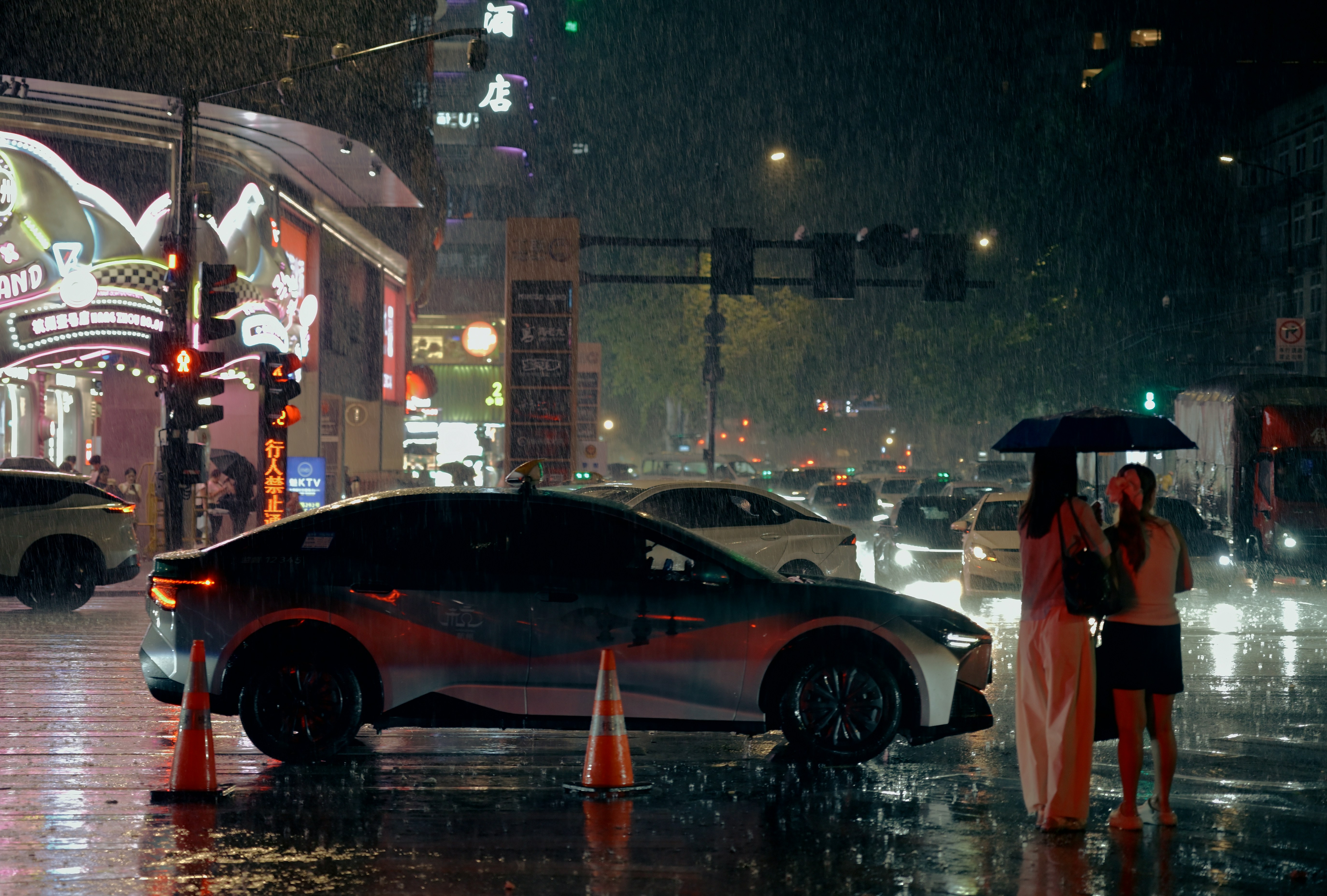 Night street scene with a dark car stopped among traffic cones; two people under an umbrella share a moment in the rain, illuminated by neon signs and reflections on the wet pavement.