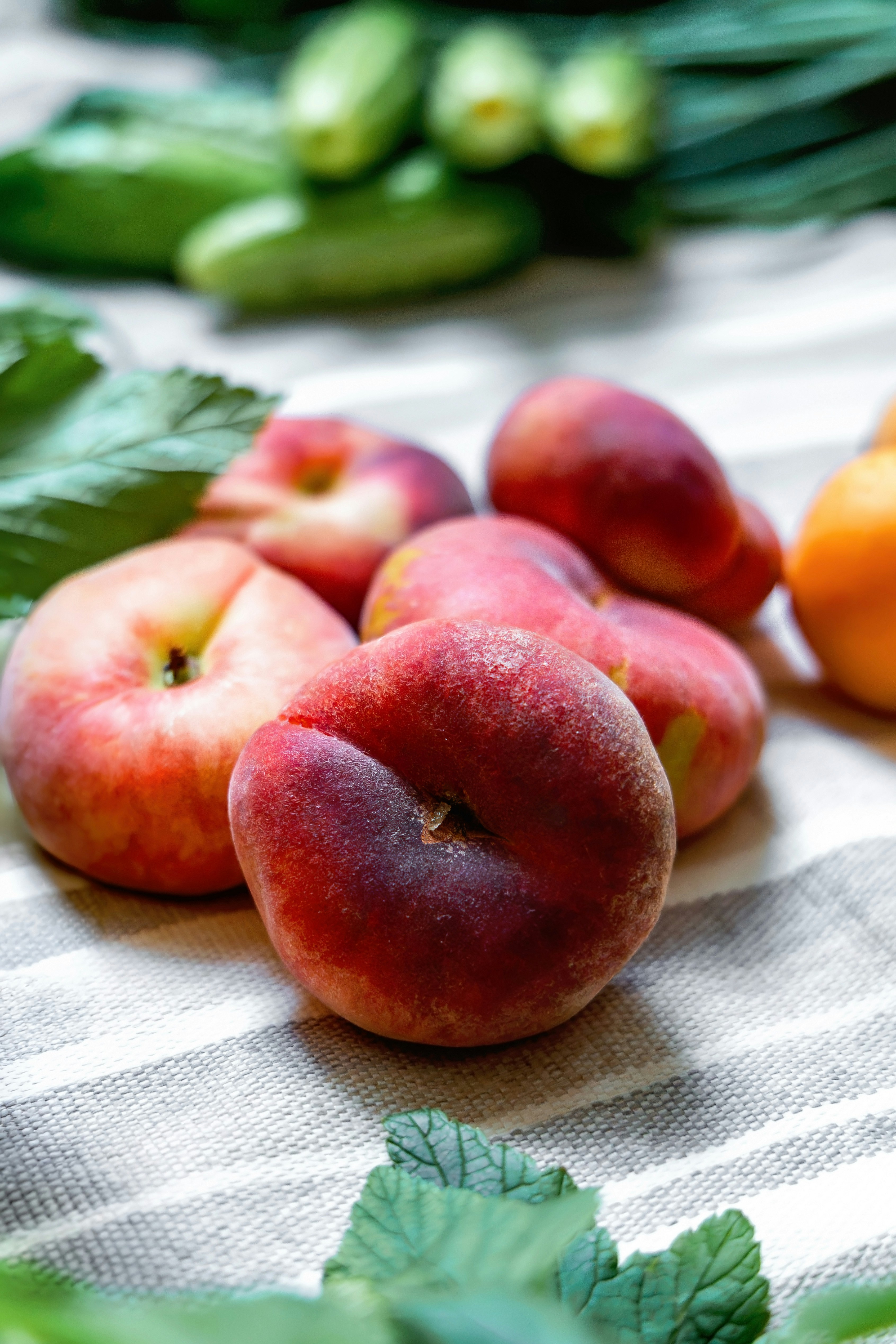 Juicy peaches rest on a striped linen backdrop with leafy greens and distant green vegetables, capturing a sunlit market-style still life.