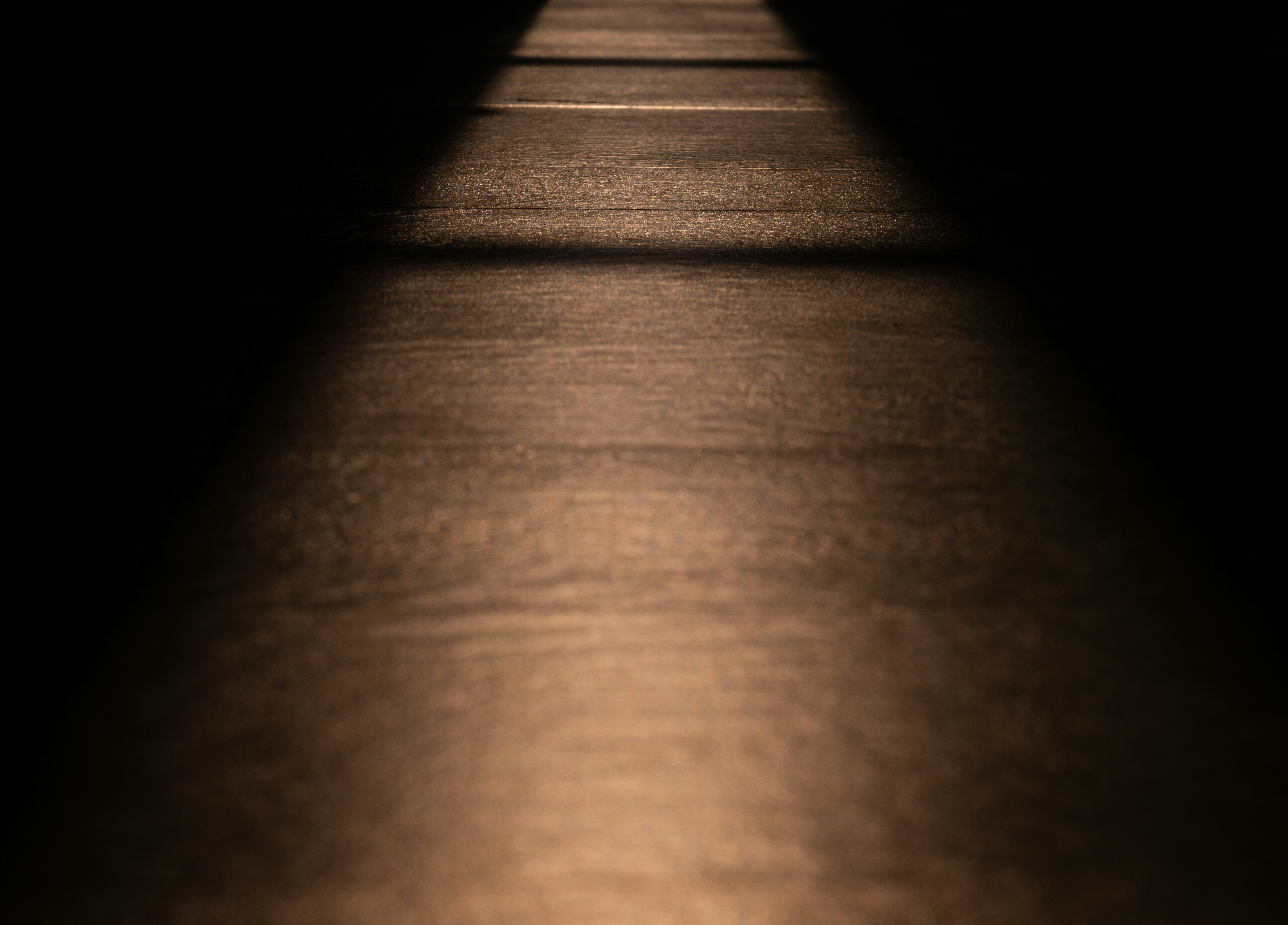 A narrow wooden floor stretches into a softly lit corridor, with warm light highlighting the texture of the planks while dark shadows frame the sides. The image feels like a deliberate study of line and tone, with the grain of the wood emphasized as a path toward the glow.