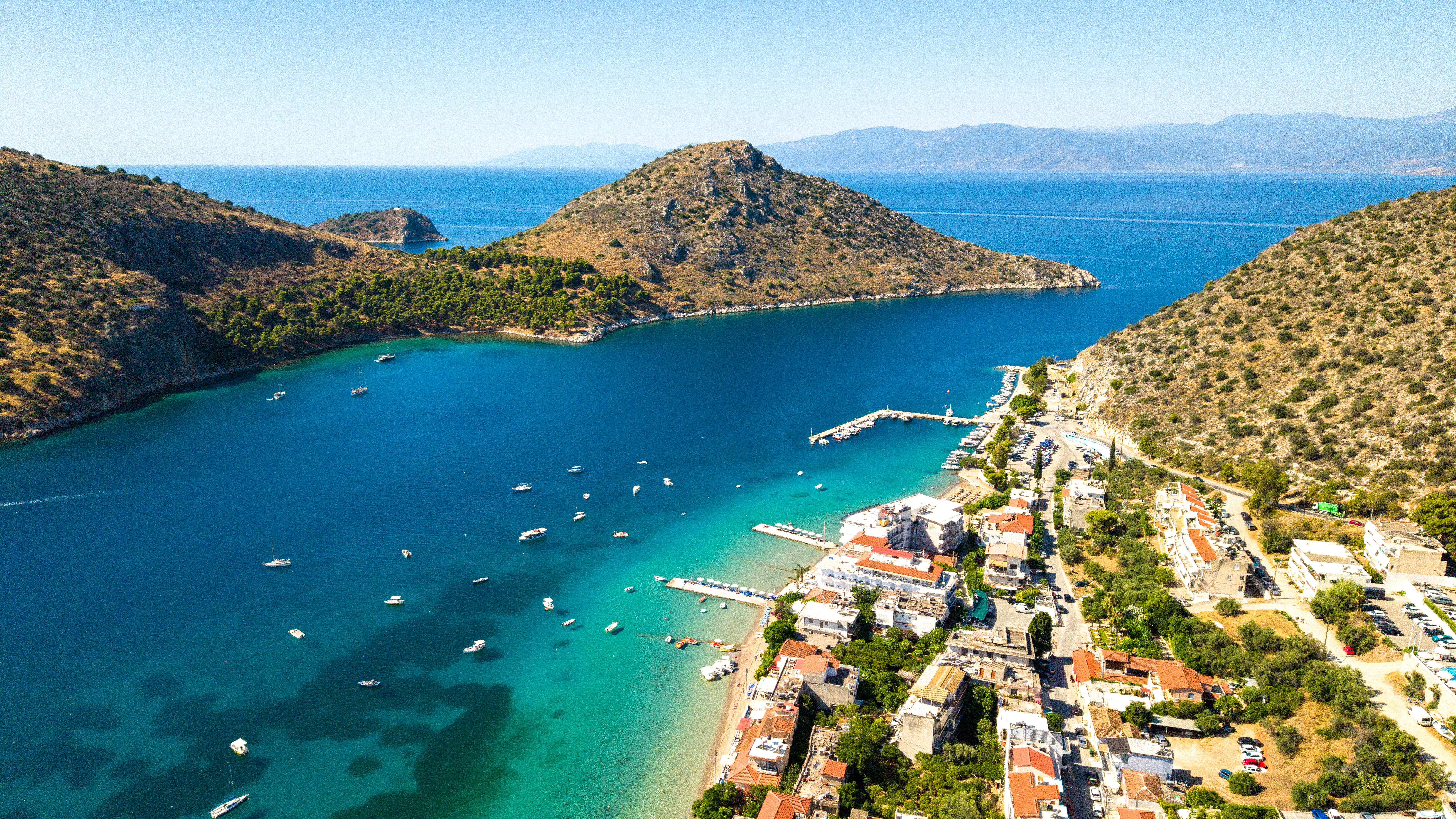 A coastal village nestles along a turquoise bay bordered by rugged hills and a white-sand beach. Docks and boats fringe the water, with the hills framing a winding shoreline and clear blue sea.