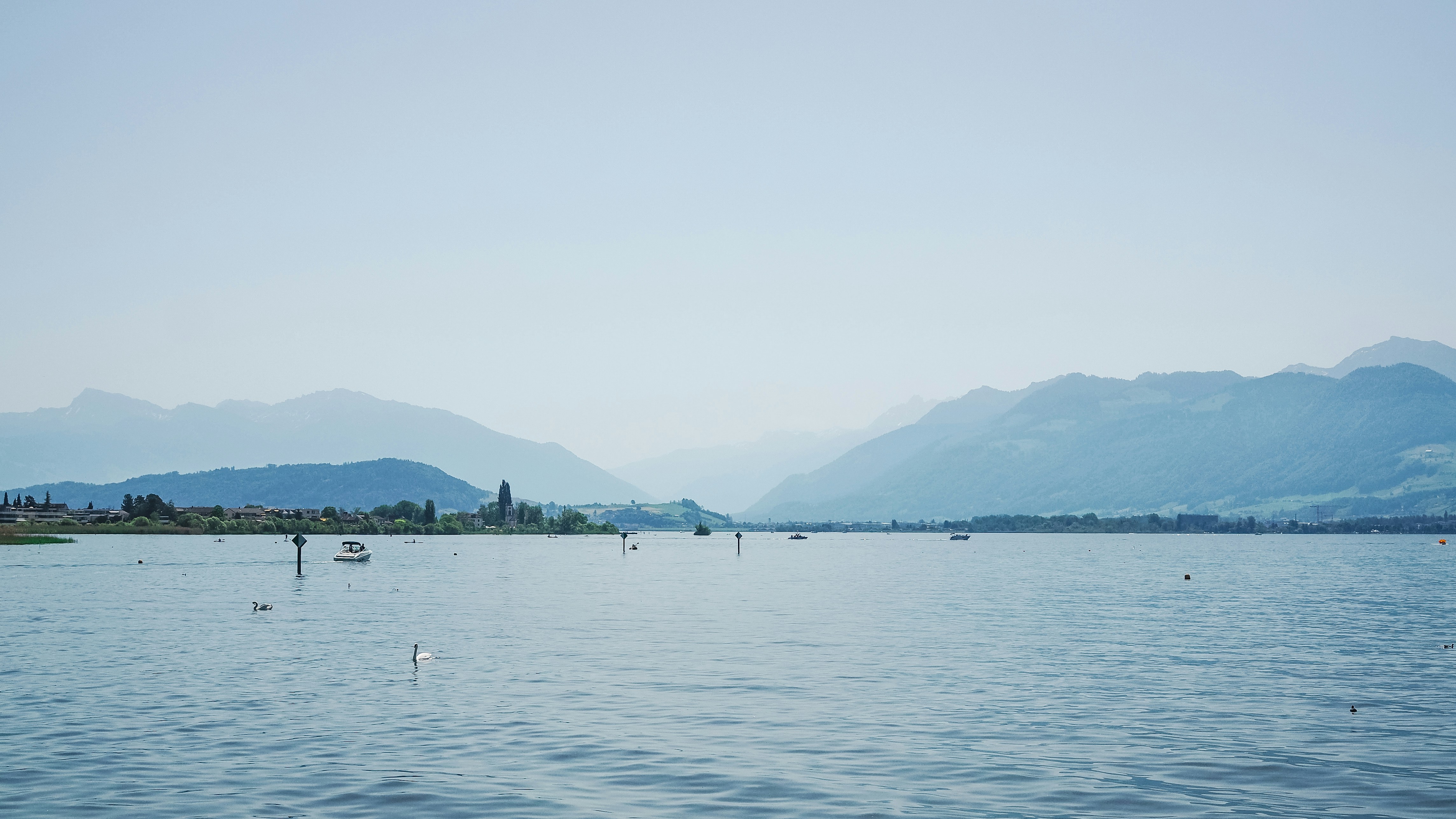 Open lake with distant mountains under a clear sky; a tranquil shoreline scene capturing calm water and faint boats in the distance. (Still image, landscape photograph.)