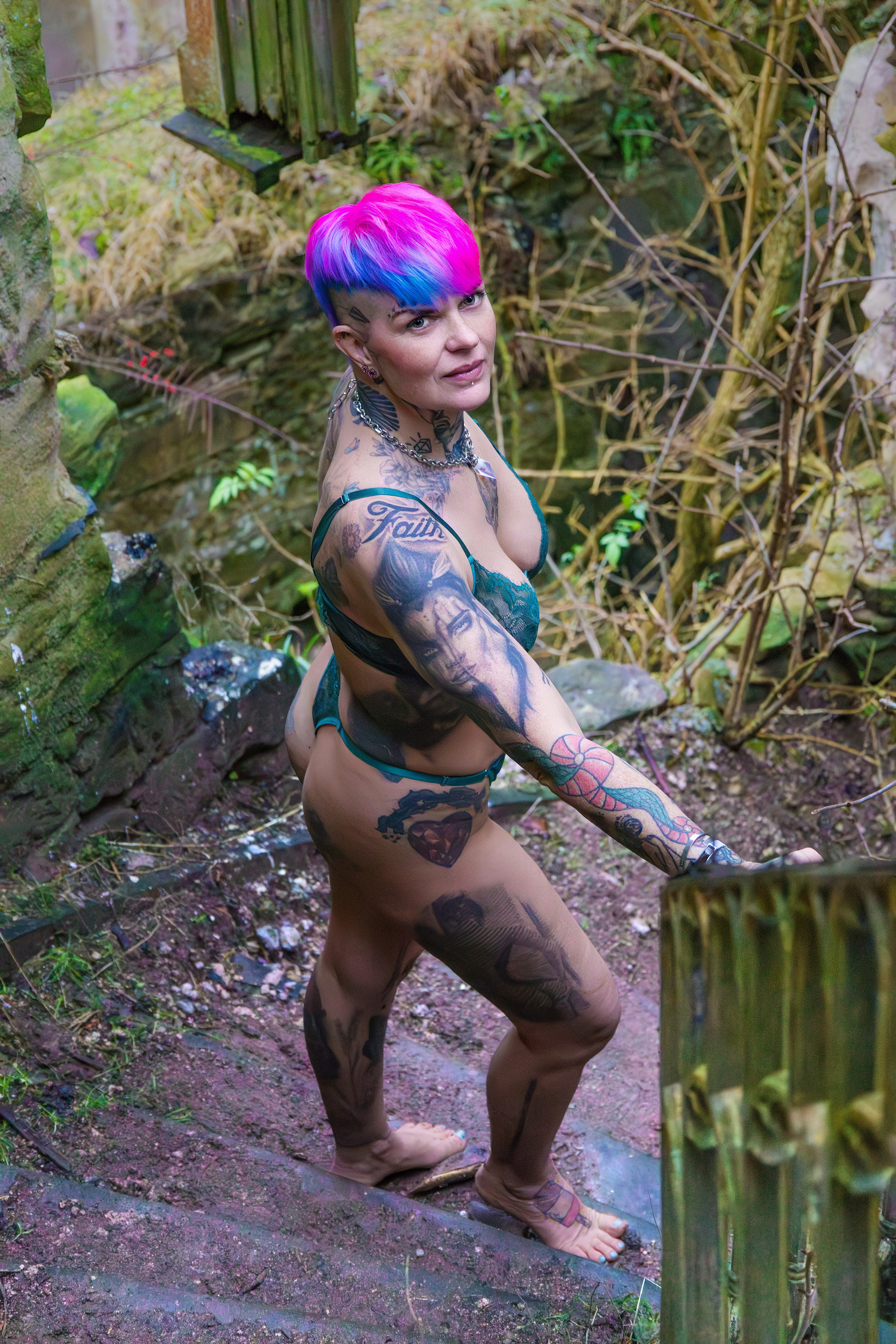 Tattooed woman in teal lingerie stands on a mossy stair beside a railing in a forested ruin, her vibrant pink-blue hair drawing focus as she gazes upward.