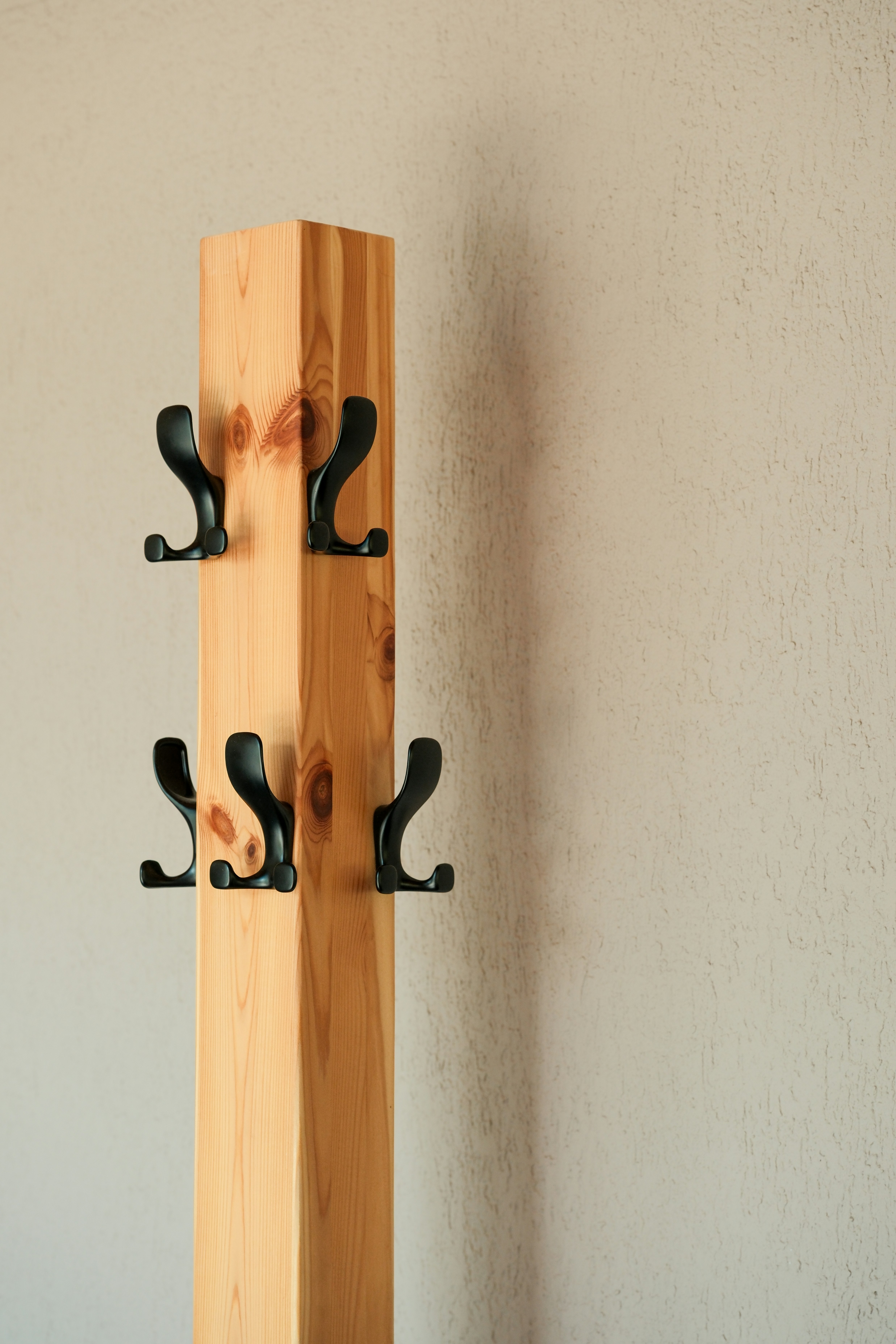Wooden coat rack with multiple black hooks stands against a textured beige wall, bathed in natural light.