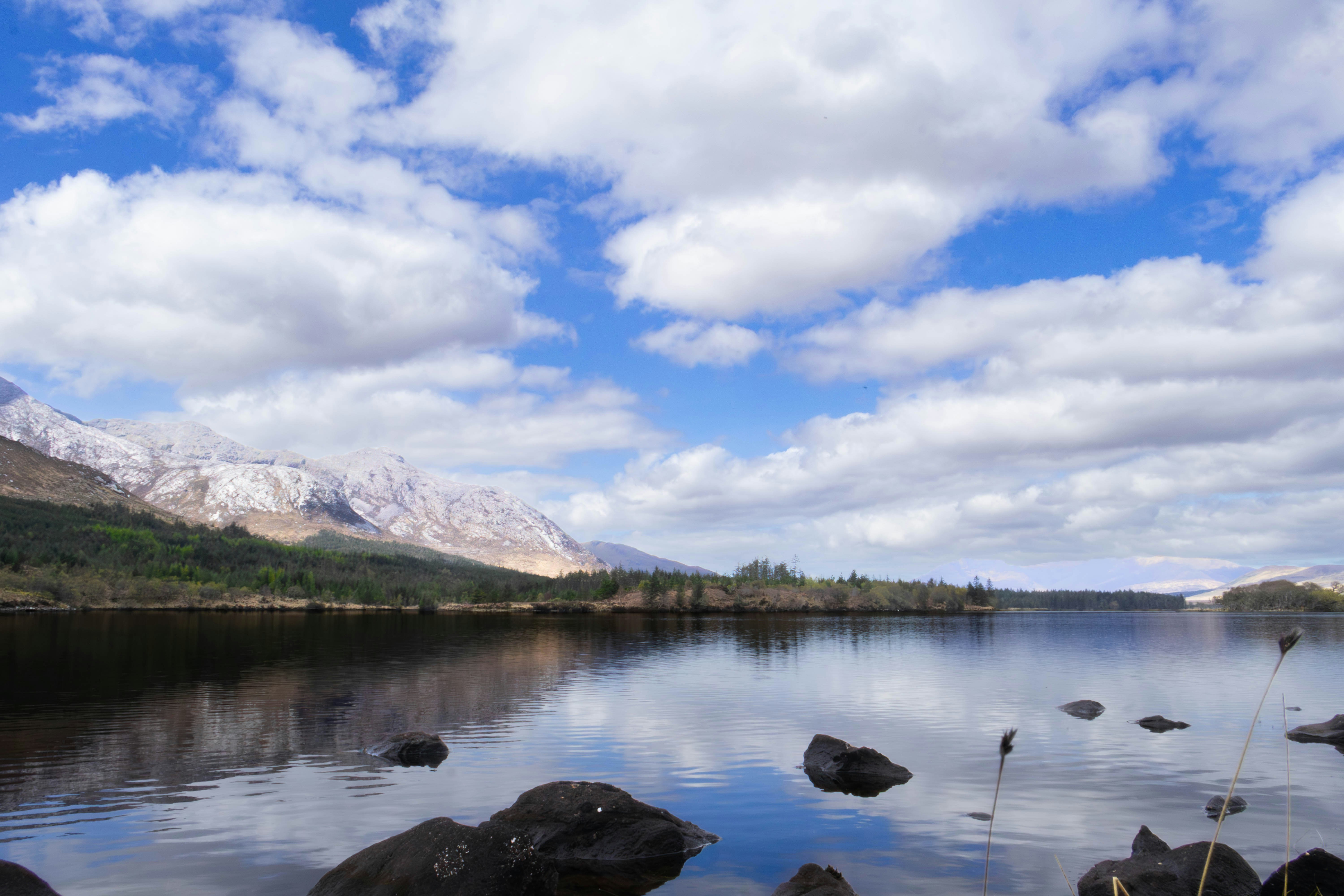 Serene alpine lake scene with rocky shore, calm water, and snow-dusted mountains under a partly cloudy sky.