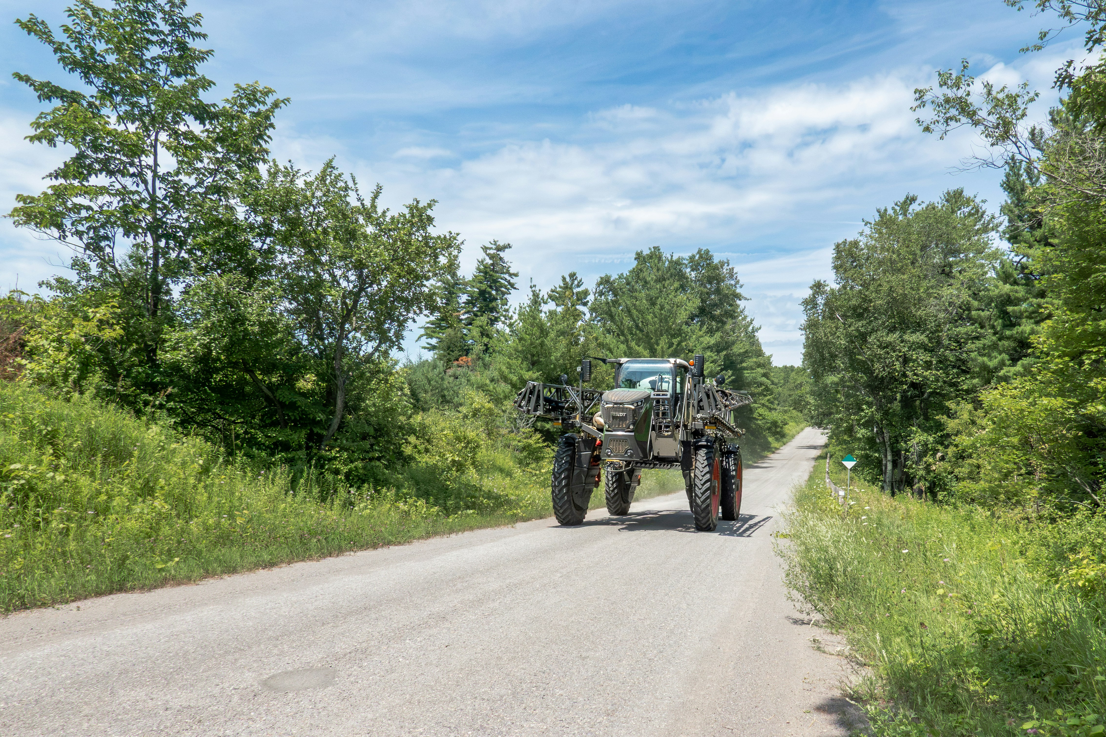 Compact all-terrain vehicle traversing a sunlit country road flanked by greenery; a rugged machine dominates the frame, with a clear sky above. This is a photo capturing the vehicle in motion on a rural route.