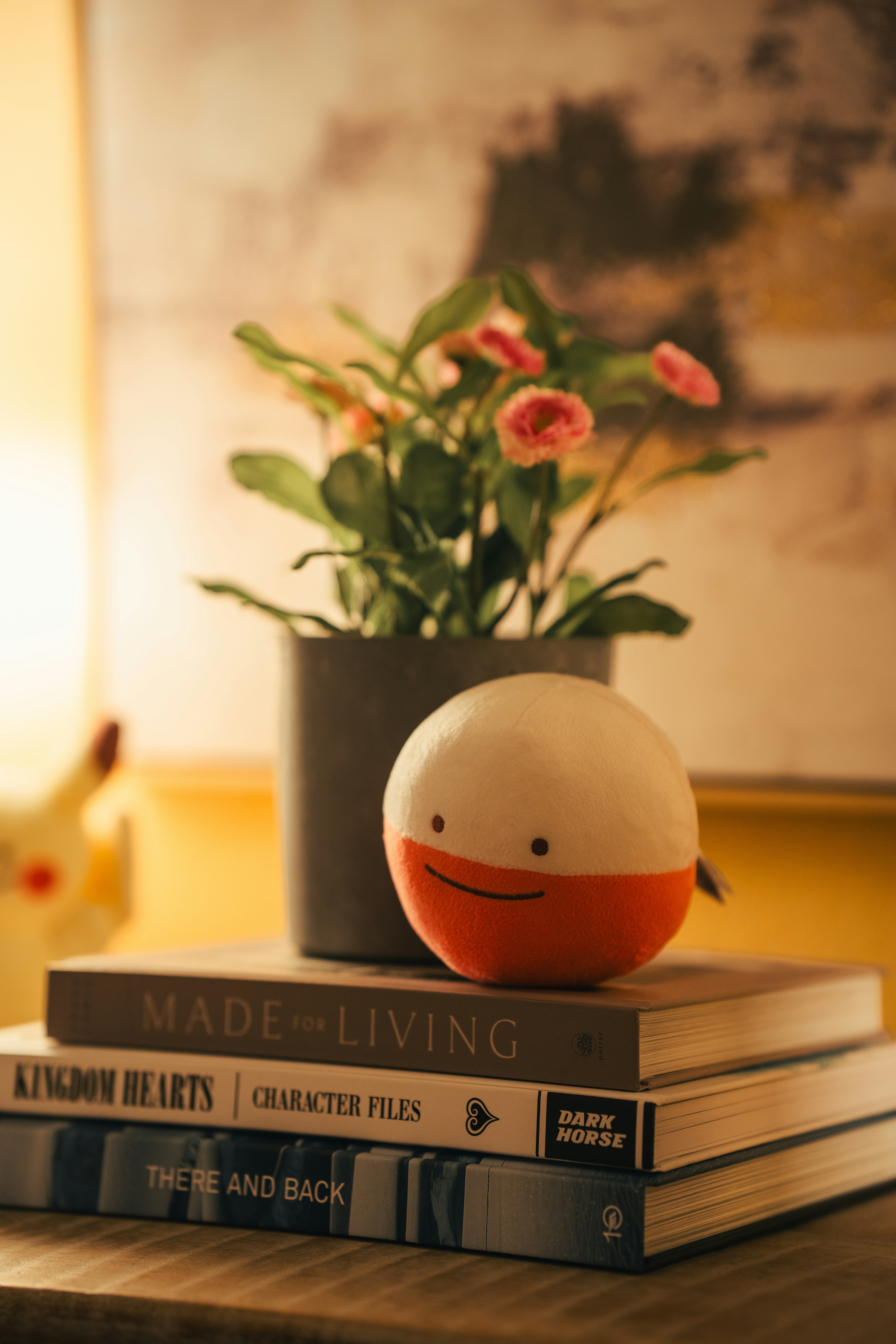 Smiling Plush orb perched atop a stack of books beside a potted bouquet