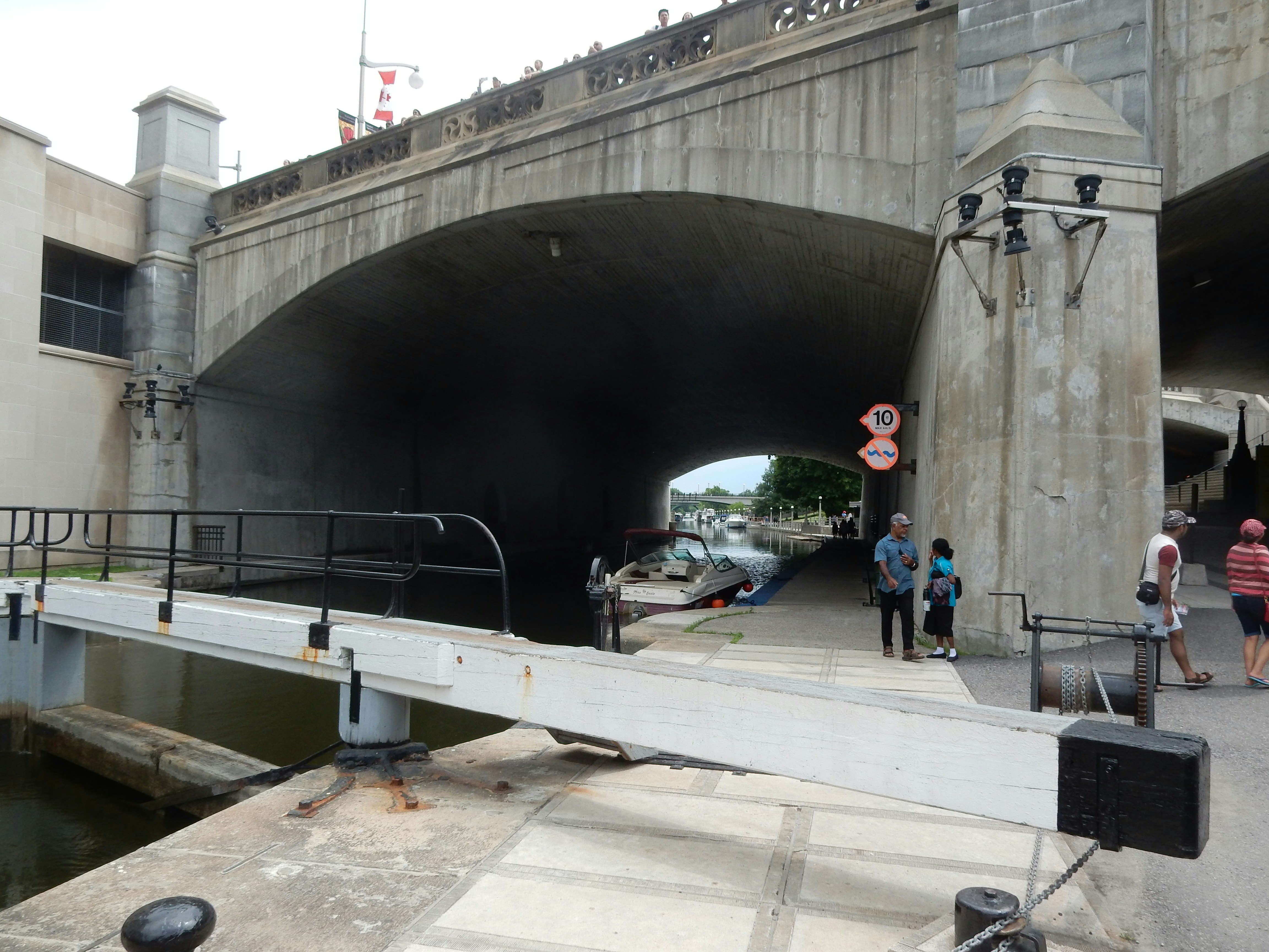 A riverside scene under a concrete bridge with a pedestrian walkway, a small boat, and a few people standing near the canal edge; a drawbridge-like structure spans the water. The image captures urban infrastructure and casual activity along the waterway.
