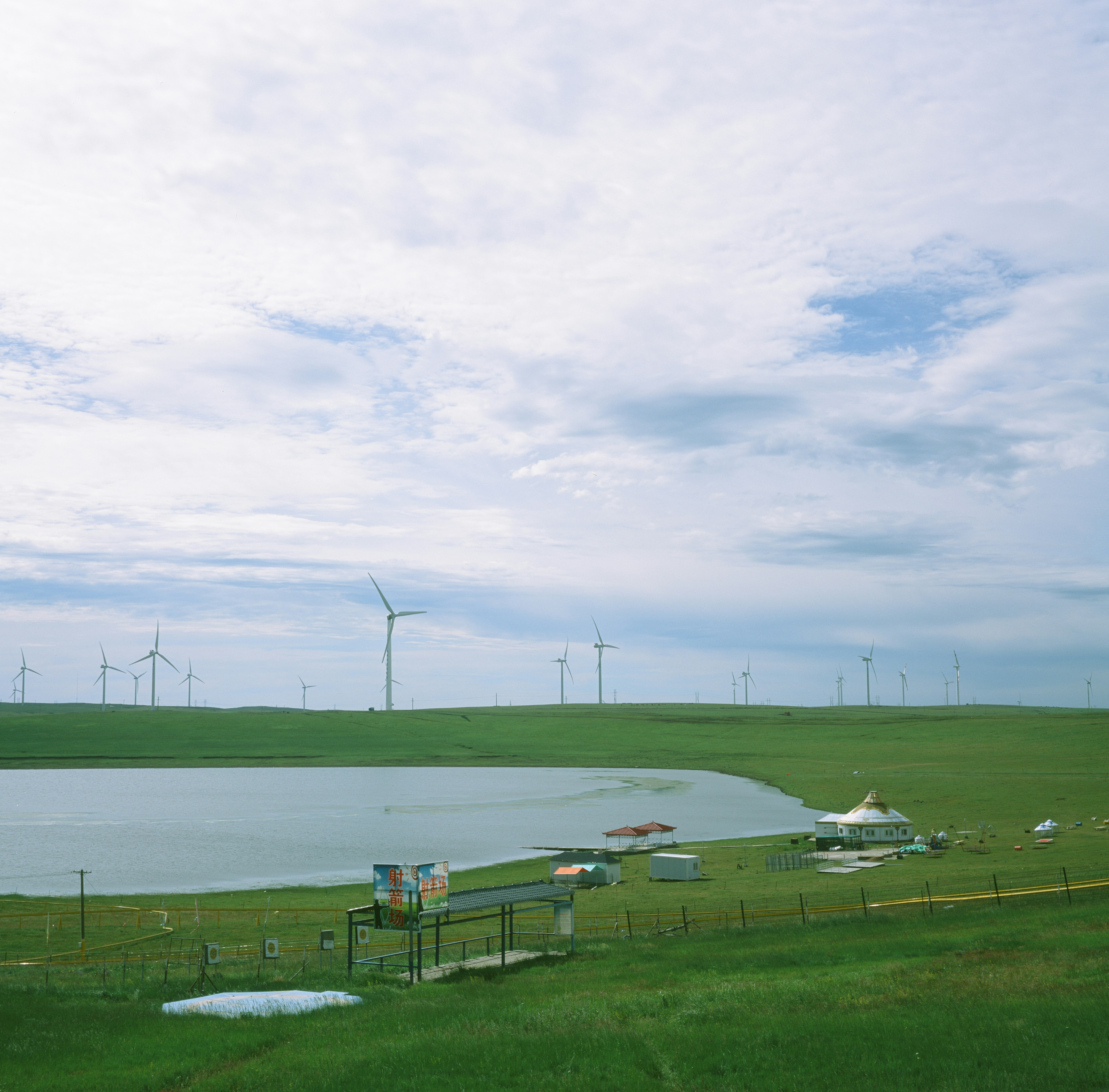 Wind turbines dot a broad green plain beside a calm lake, with a small cluster of buildings and a fence in the foreground under a soft, expansive sky.