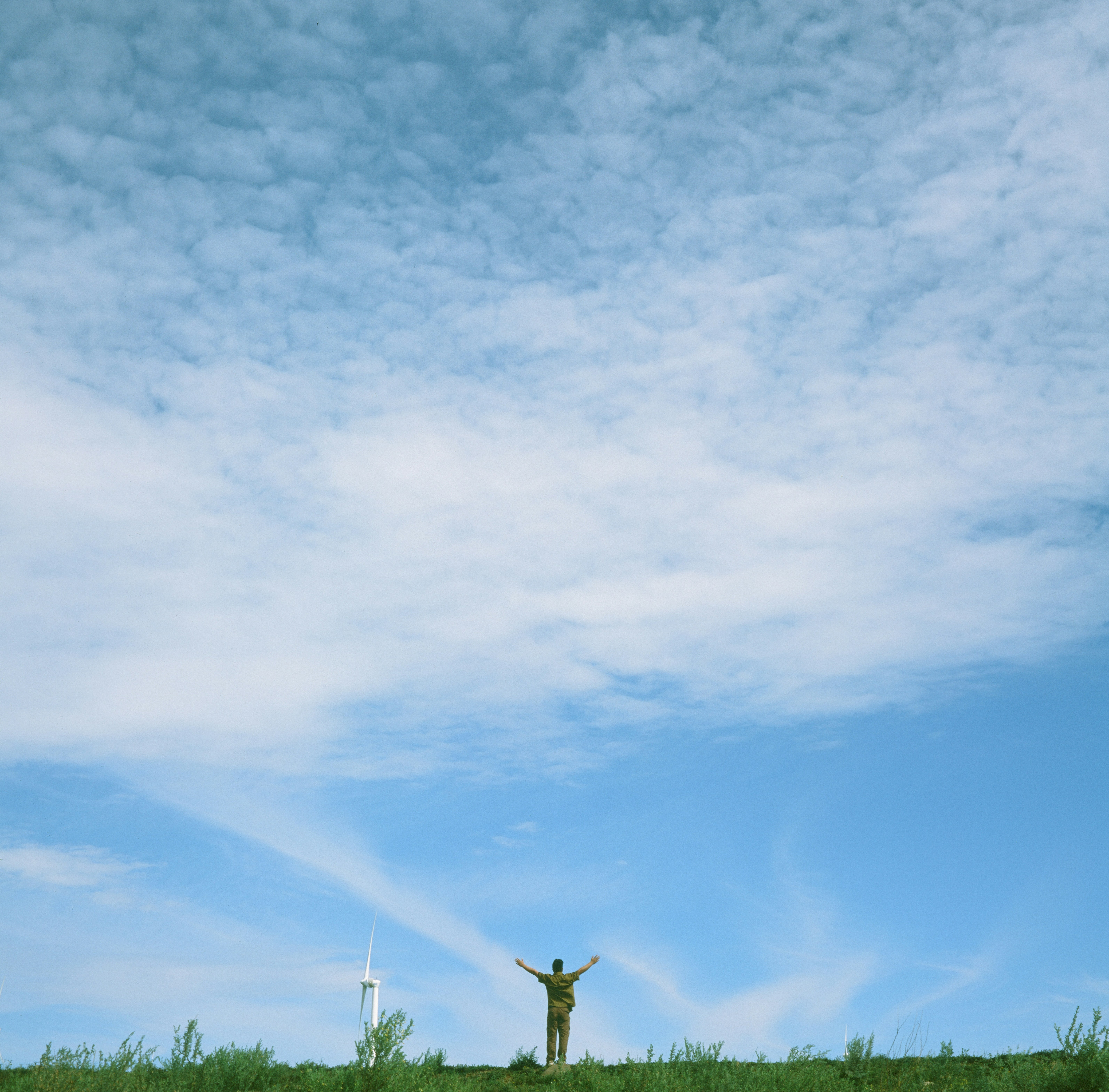 A lone person stands on a grassy ledge with arms raised against a vast blue sky and wispy clouds; a wind turbine is visible to the left, suggesting a landscape scene.