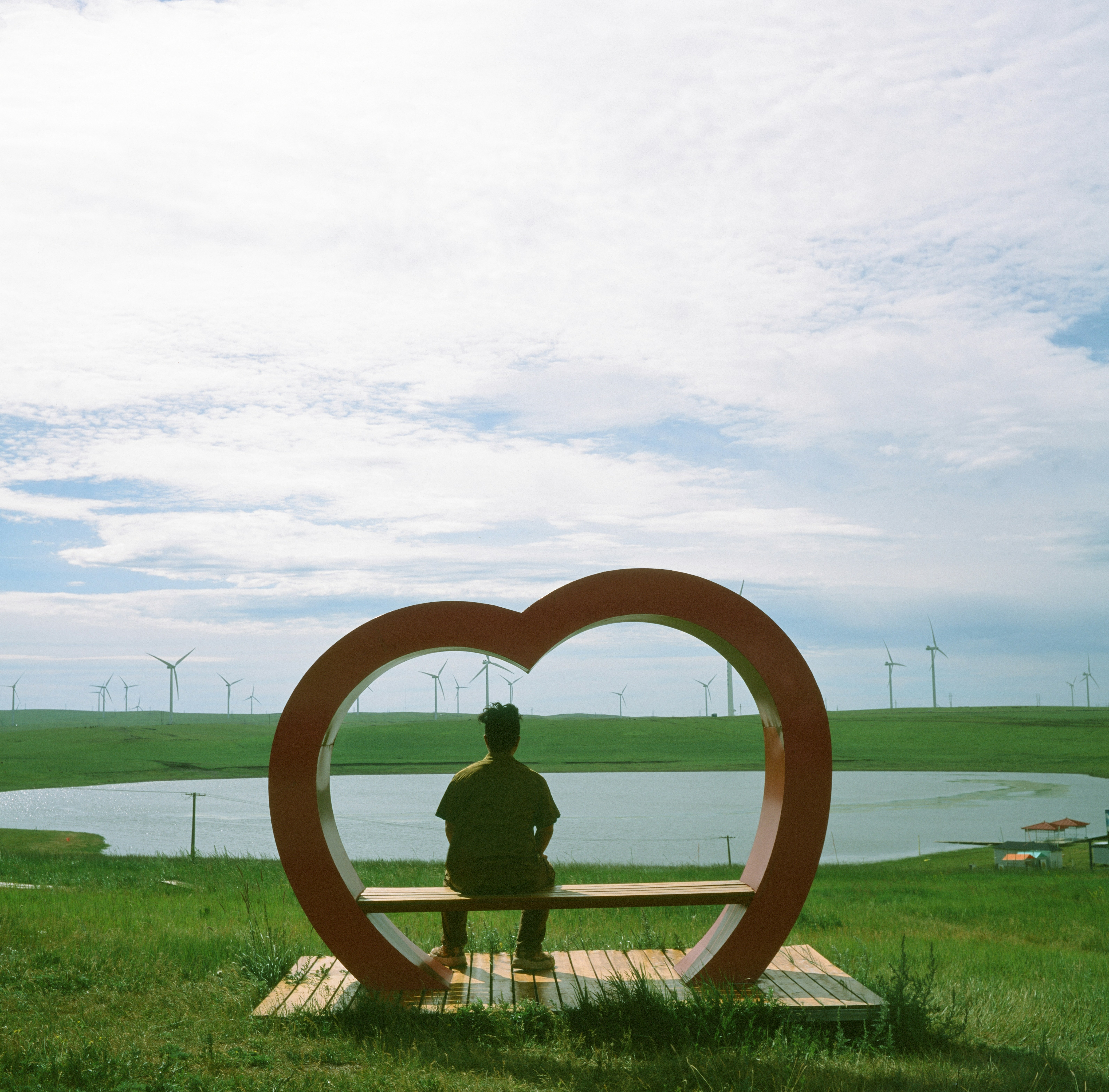 Person seated on a wooden bench framed by a heart-shaped sculpture overlooking a wind farm and a small body of water; peaceful landscape scene with a cloudy sky.