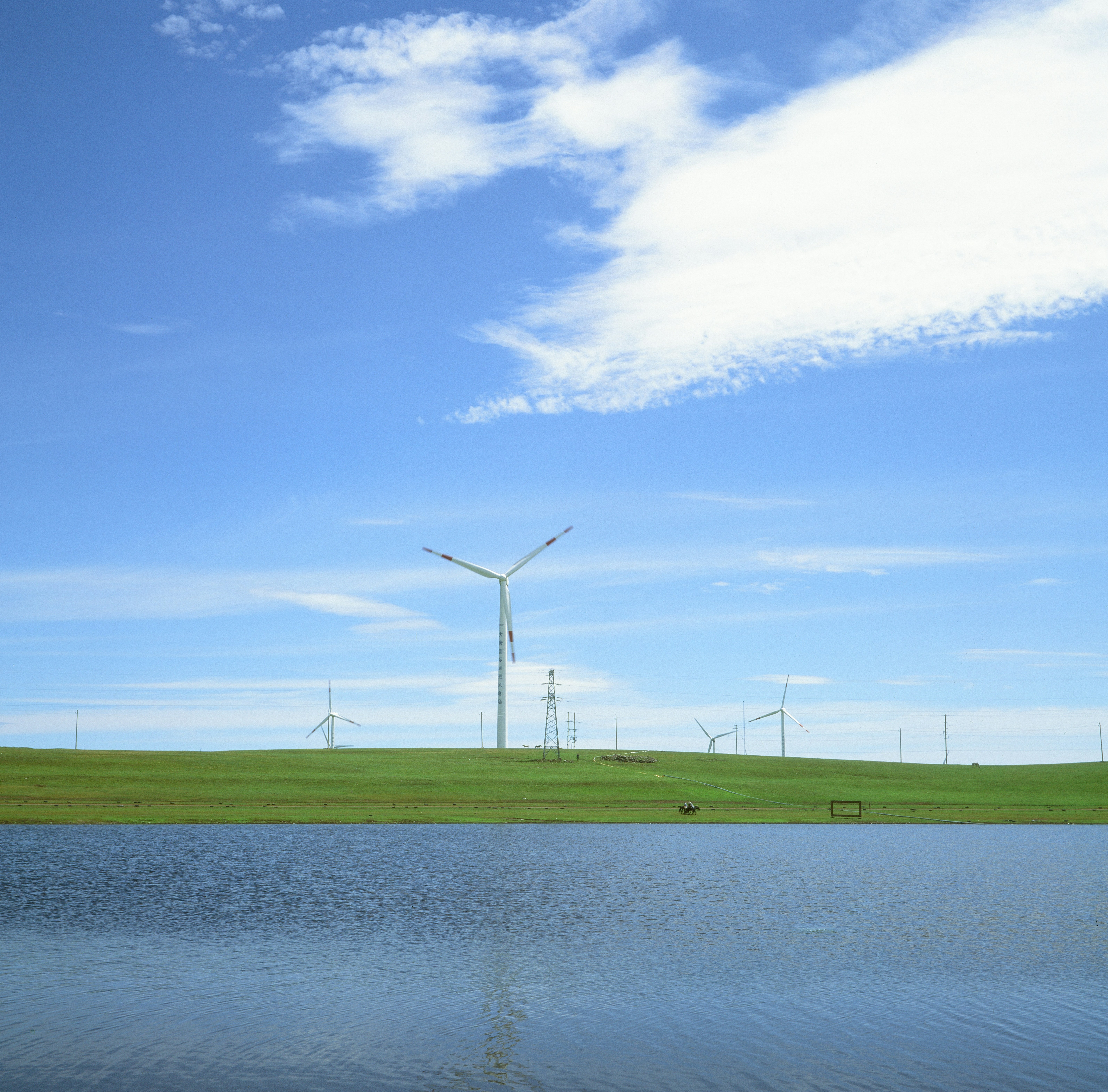 Wind turbines dot a flat green horizon beside a calm lake, under a bright blue sky. A tranquil landscape emphasizes sustainable energy quietly powering the scene.
