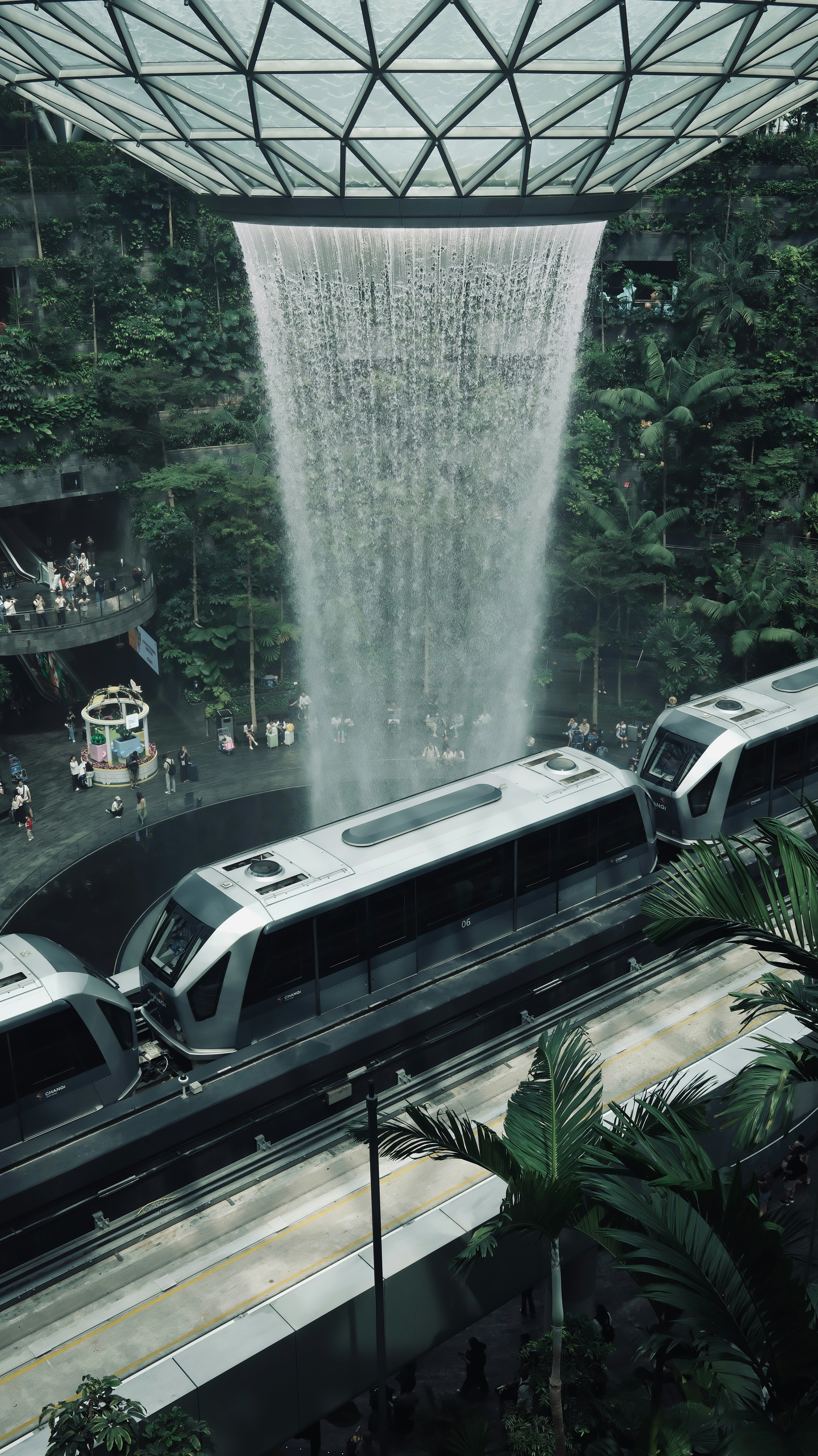 A tall indoor waterfall arcs from a glass ceiling into a lush atrium, with sleek monorail trains passing along elevated tracks and visitors gathered around the base. A subtle cool tone dominates the scene, highlighting architectural greenery.