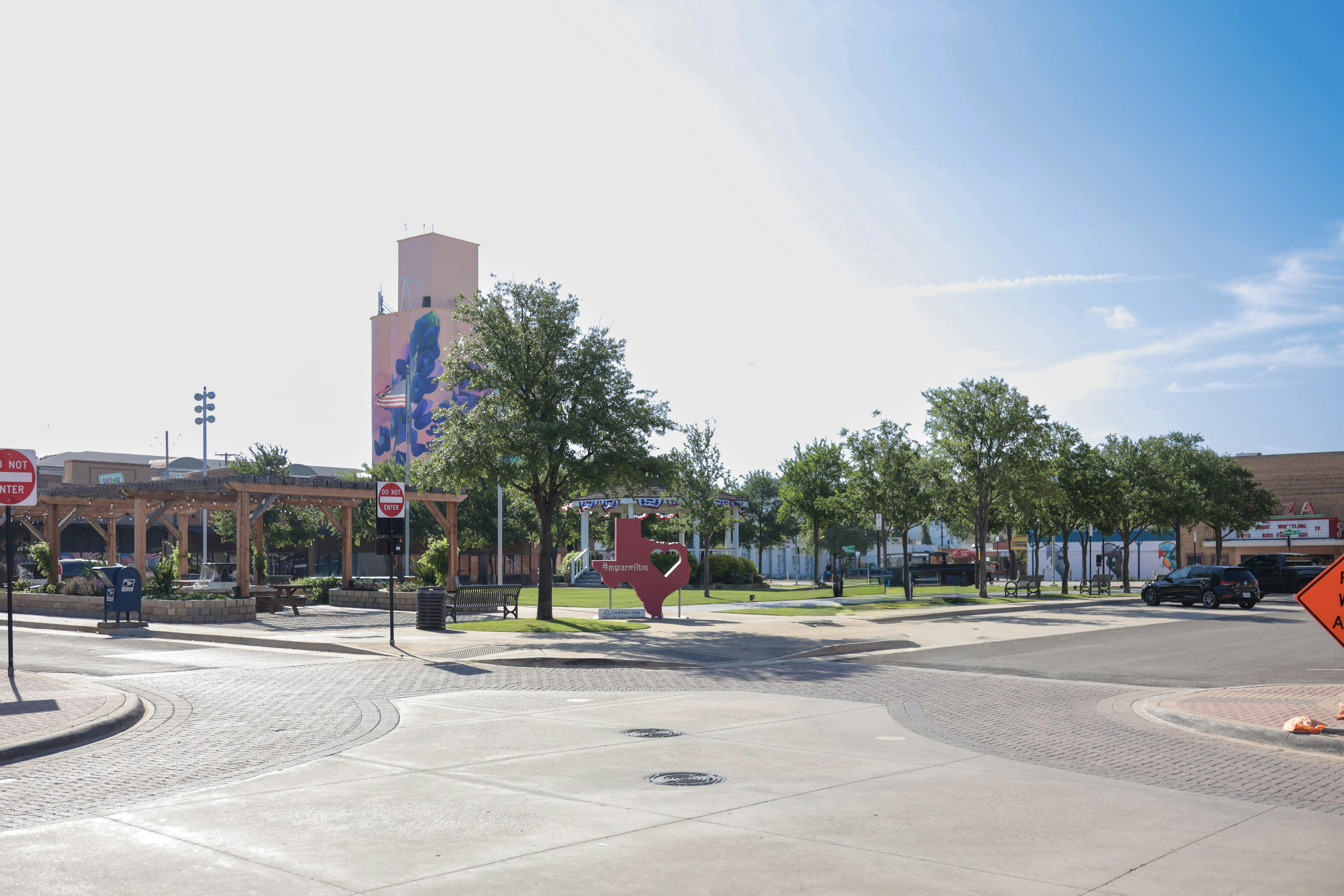 Wide plaza scene with a red sculpture and palm tree-lined backdrop in a sunny urban setting. A curved road and pedestrian area frame the sculpture, hinting at a public square in front of a low-rise civic building.