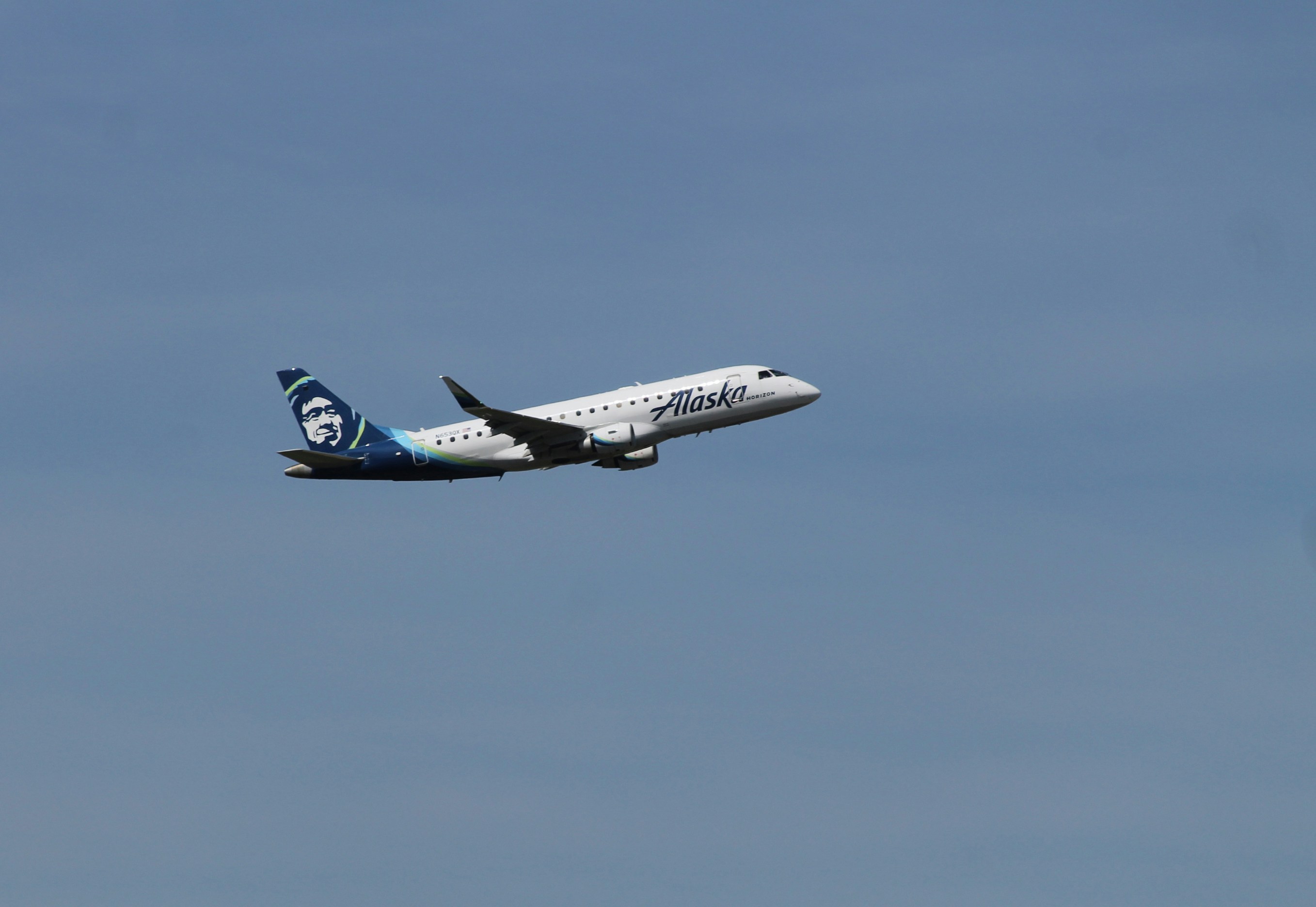 Alaska Airlines jet climbs into a clear blue sky, tail featuring the distinctive blue logo, mid-flight with slight upward angle.