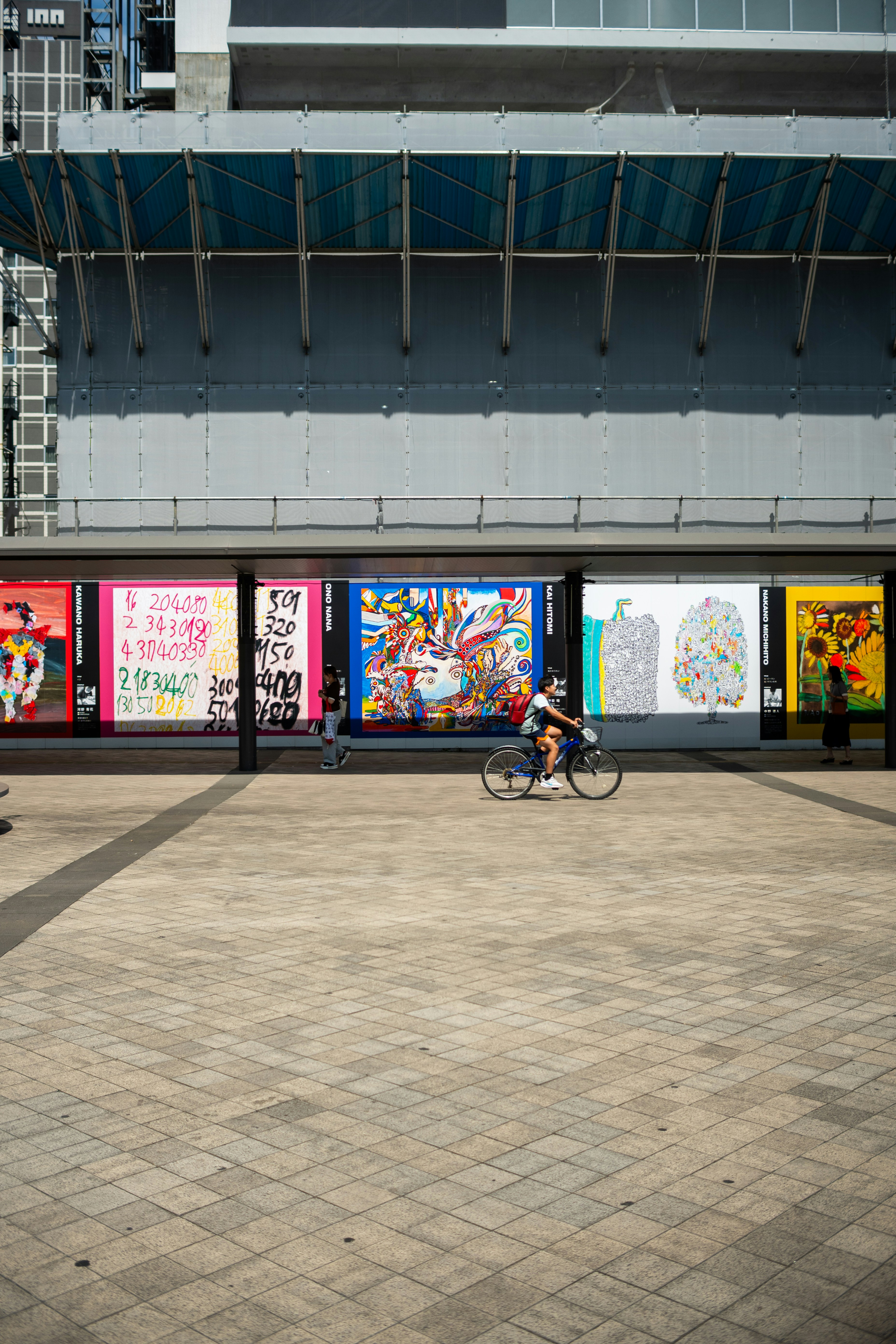 Bicyclist rides past colorful storefront murals along a modern city plaza with a stark, geometric building overhead.
