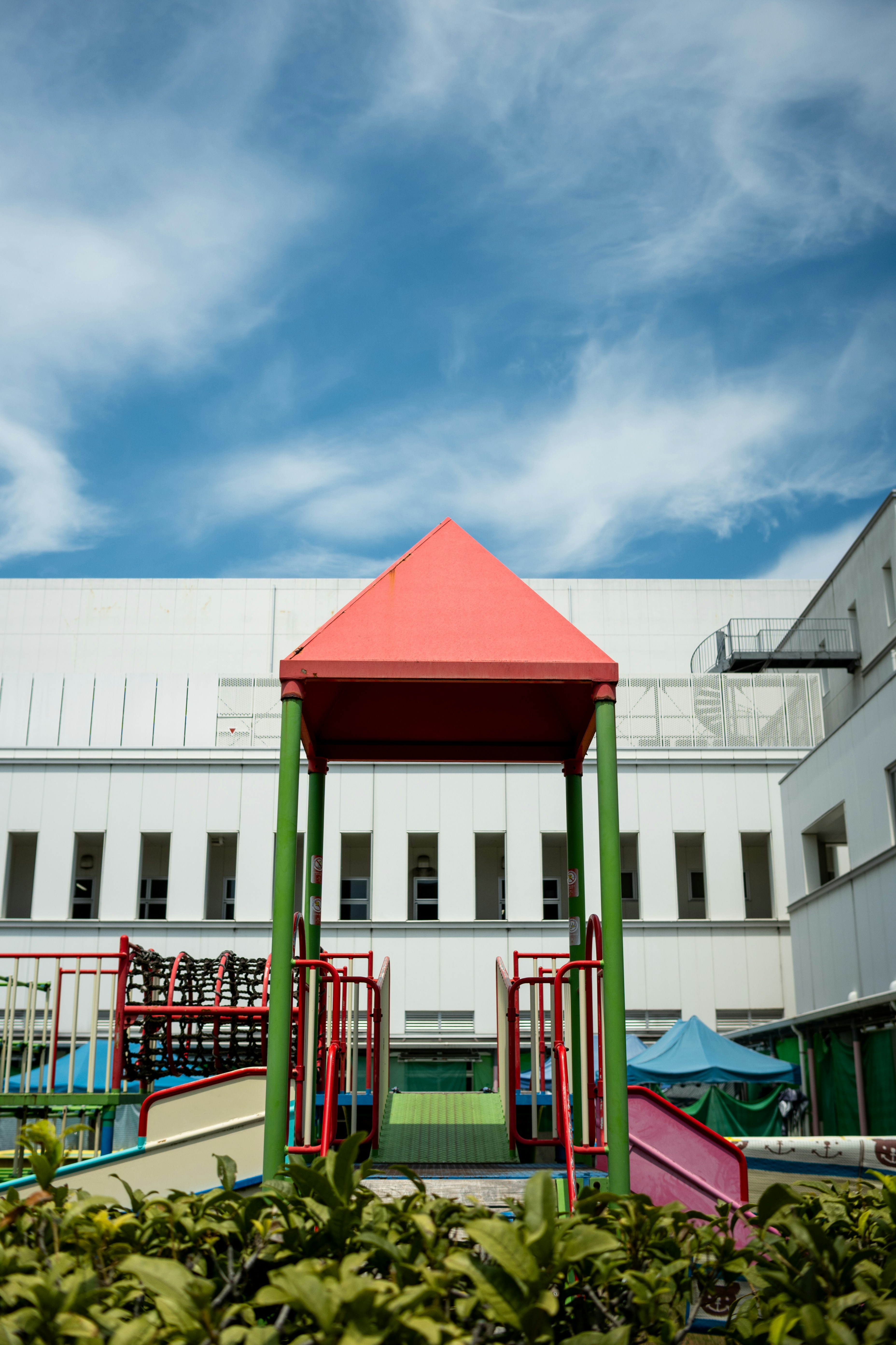 Colorful playground structure centers the scene with a red pyramid roof against a bright blue sky and white school building; foreground foliage adds depth and context to a sunny courtyard.