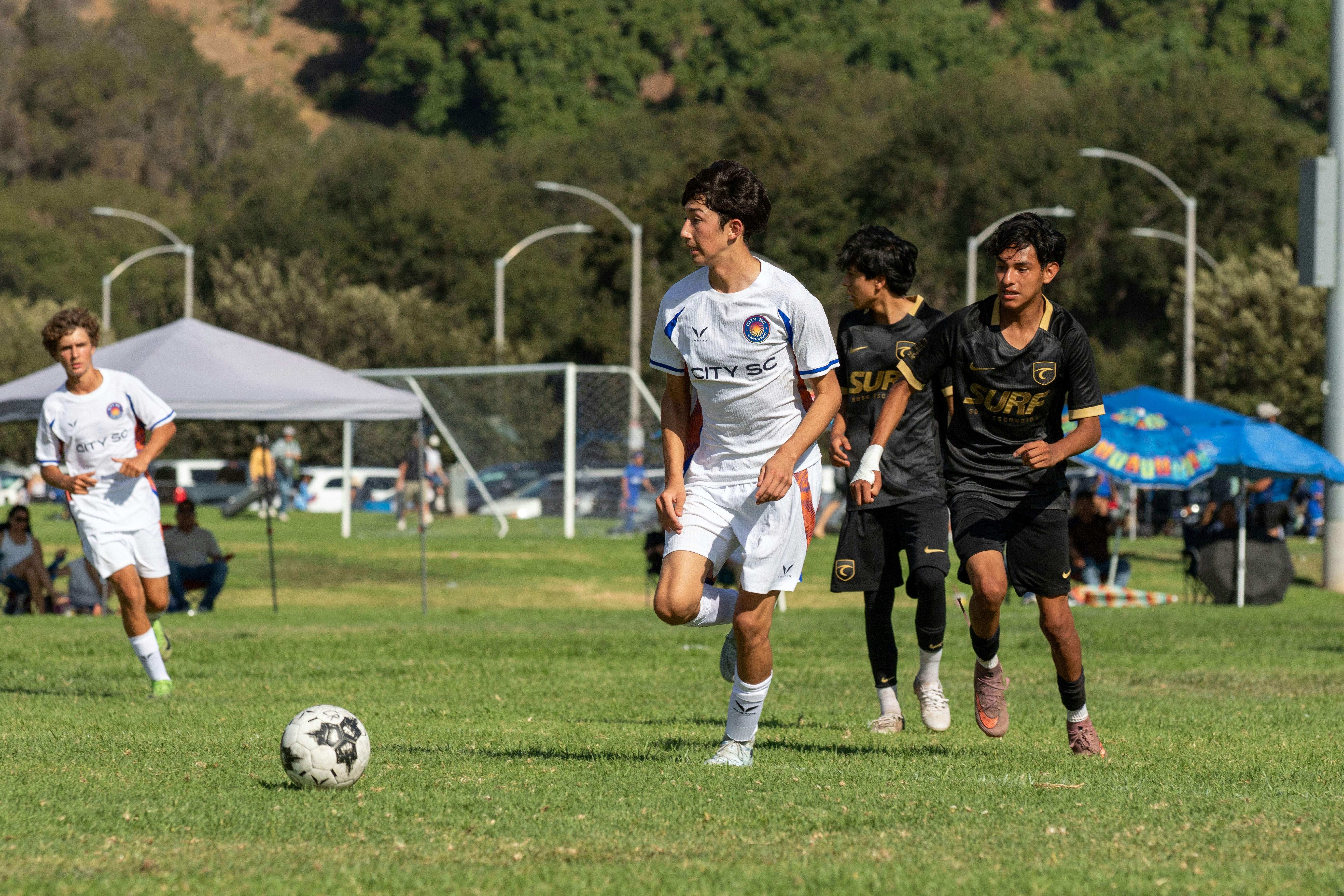 A soccer scene with a white-jersey player advancing toward a ball on a sunlit field, chased by black-jersey opponents; foreground action with tents and spectators in the background. This is a sports photograph.