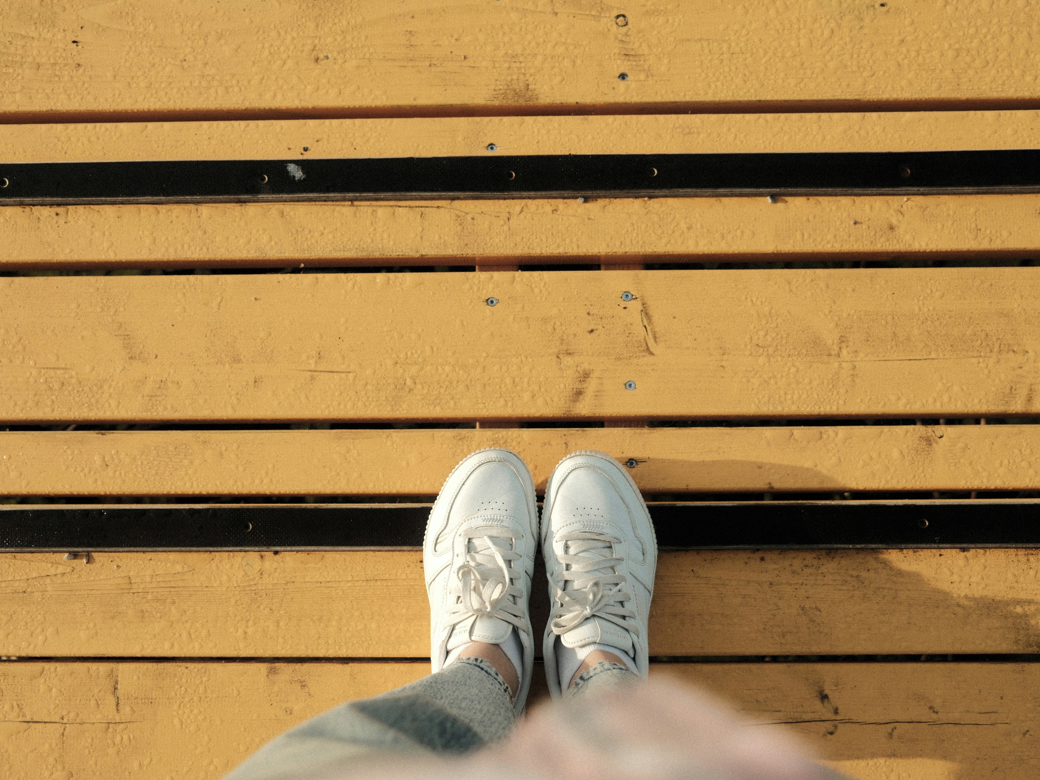 Two white sneakers stand on yellow wooden planks, seen from a seated perspective; a casual, pedestrian moment captured on a boardwalk surface.