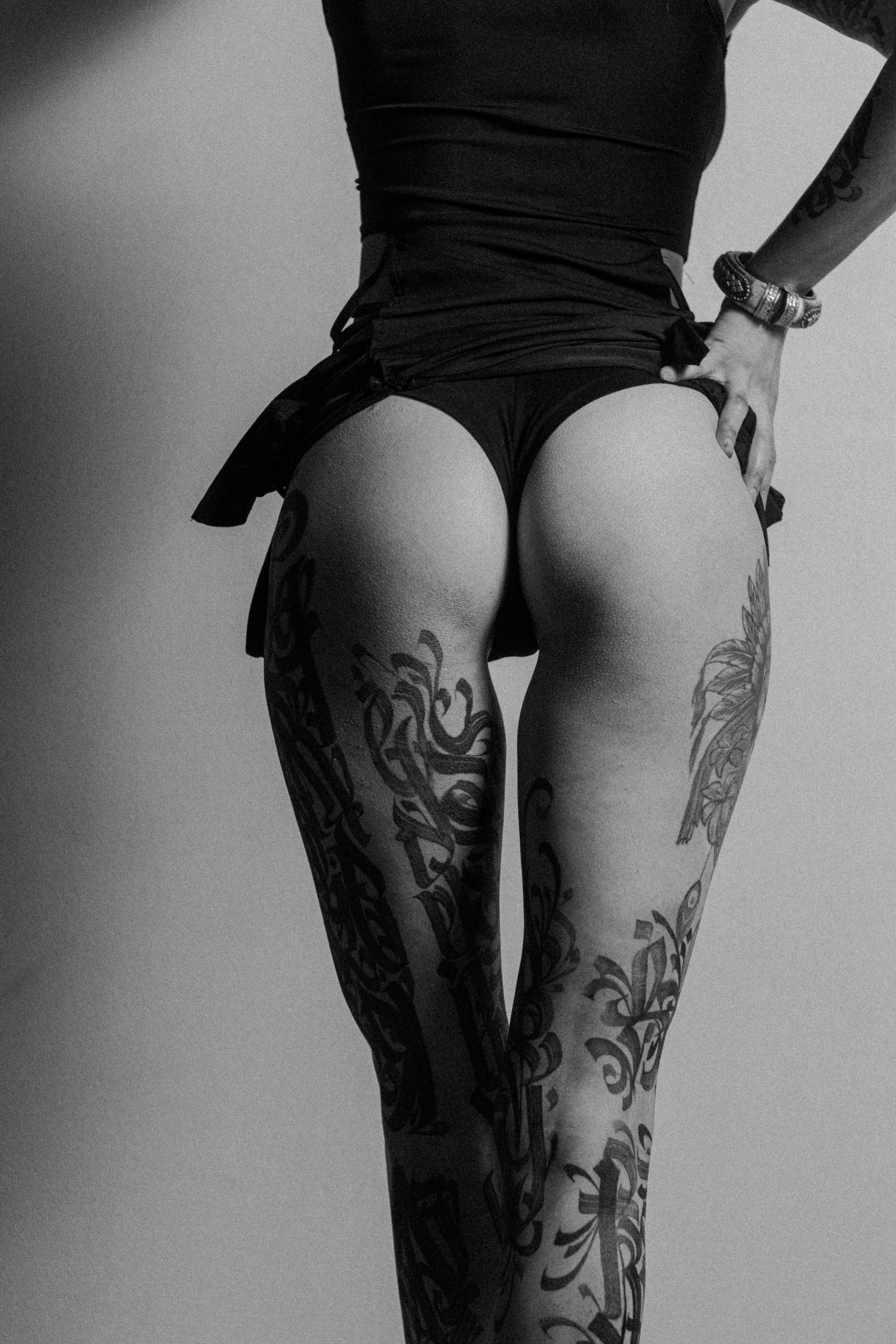 Monochrome portrait focusing on sculpted legs with extensive tattoos, posed in high-contrast studio lighting. A garment reveals the tattoos and curves, emphasizing texture and form.
