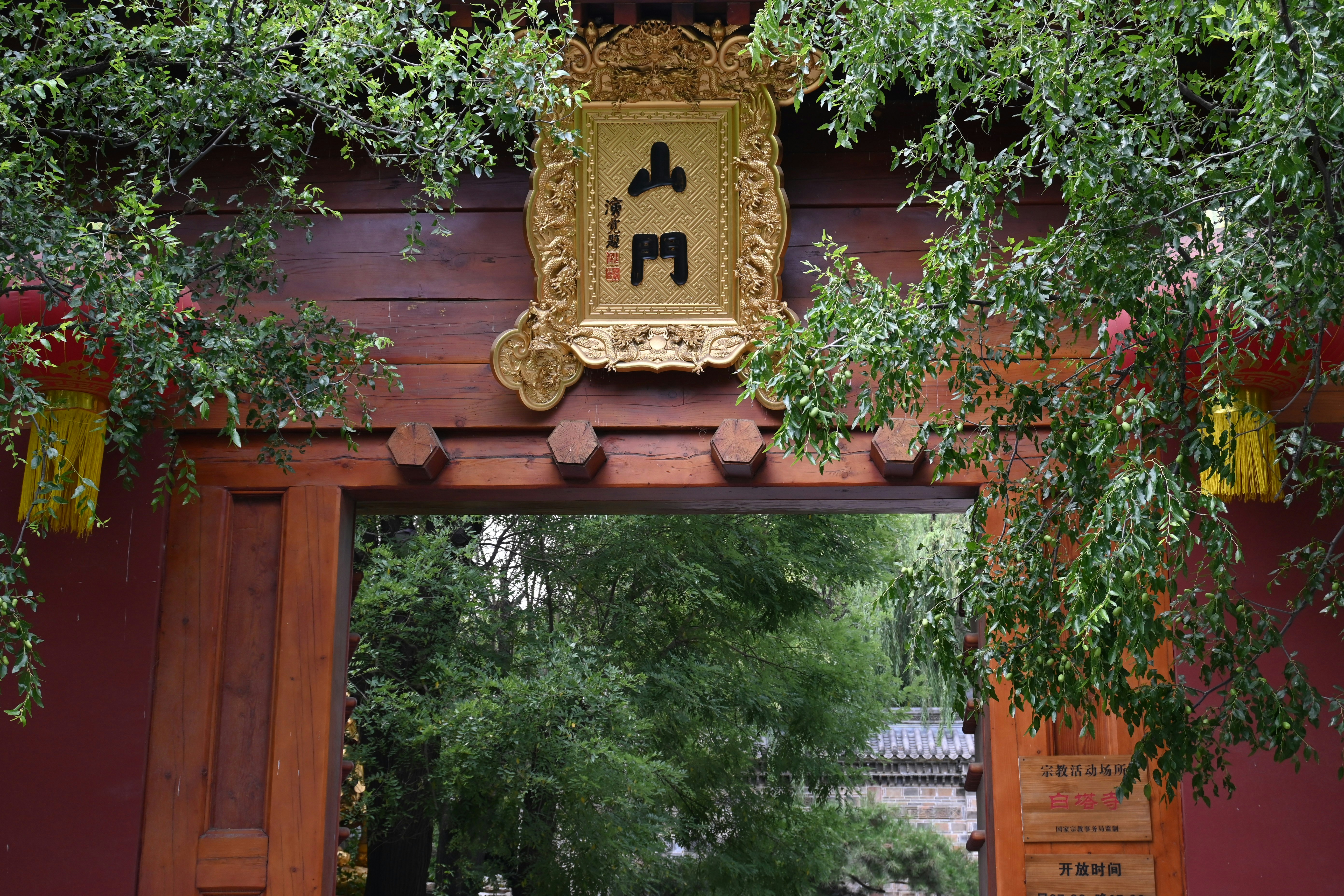 Red wooden temple gate with ornate gold plaque above and lush green foliage framing the doorway, leading into a tranquil garden. Photograph captures the traditional architecture of a Chinese temple gate.