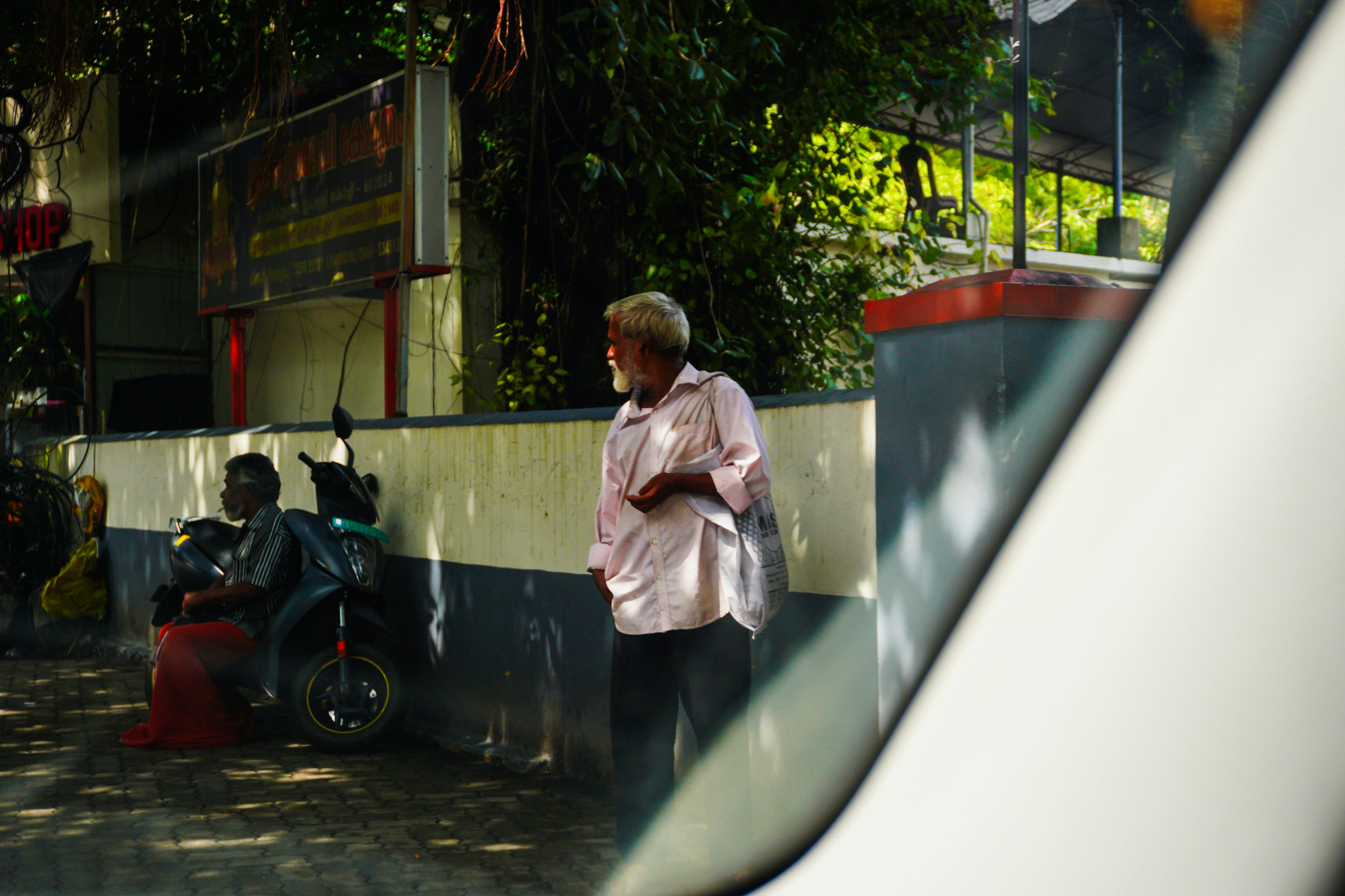 Female pedestrian in pink shirt pauses by shaded wall near scooter on a sunlit street