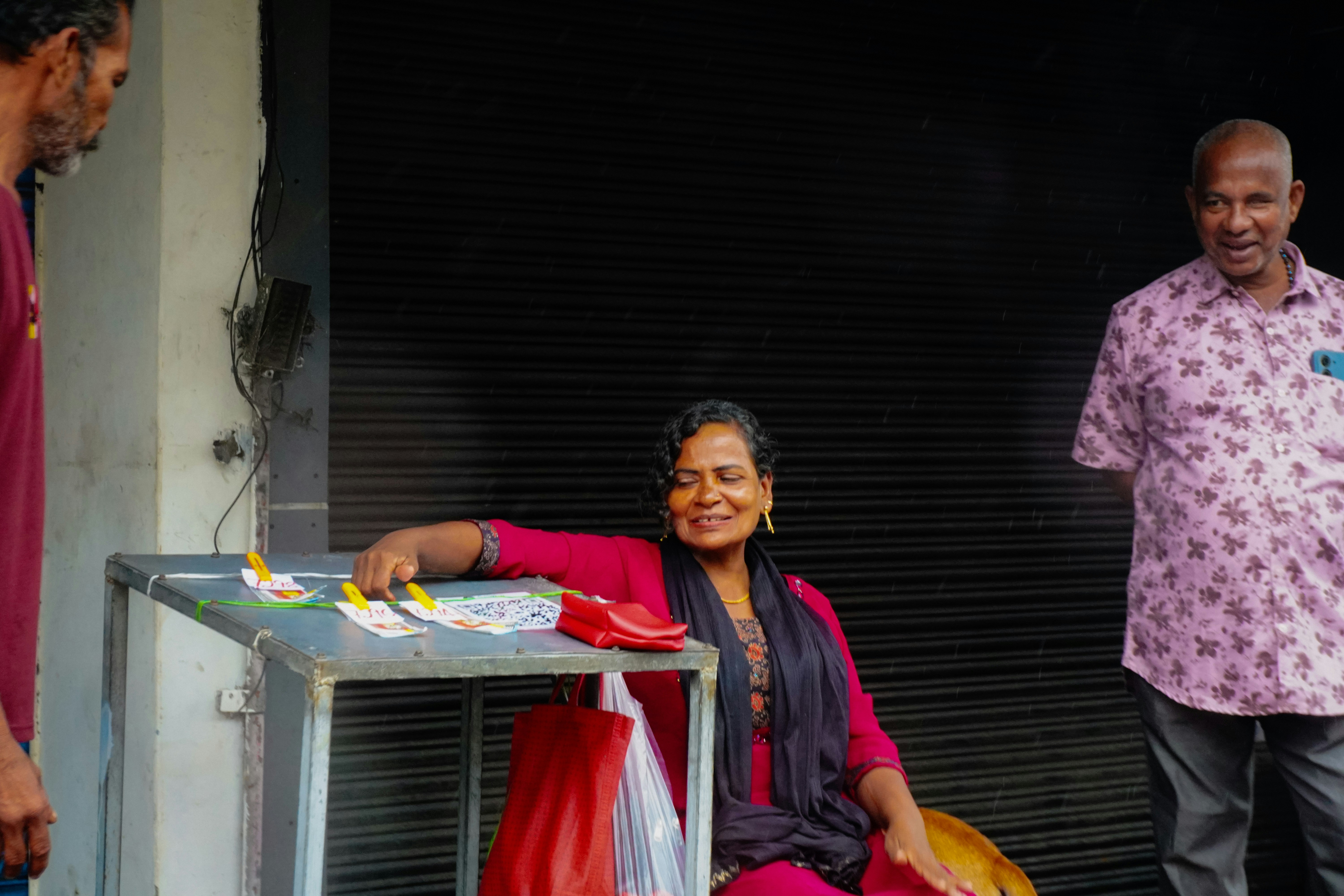 Woman in magenta shawl resting beside a street-side counter, under a shaded shopfront