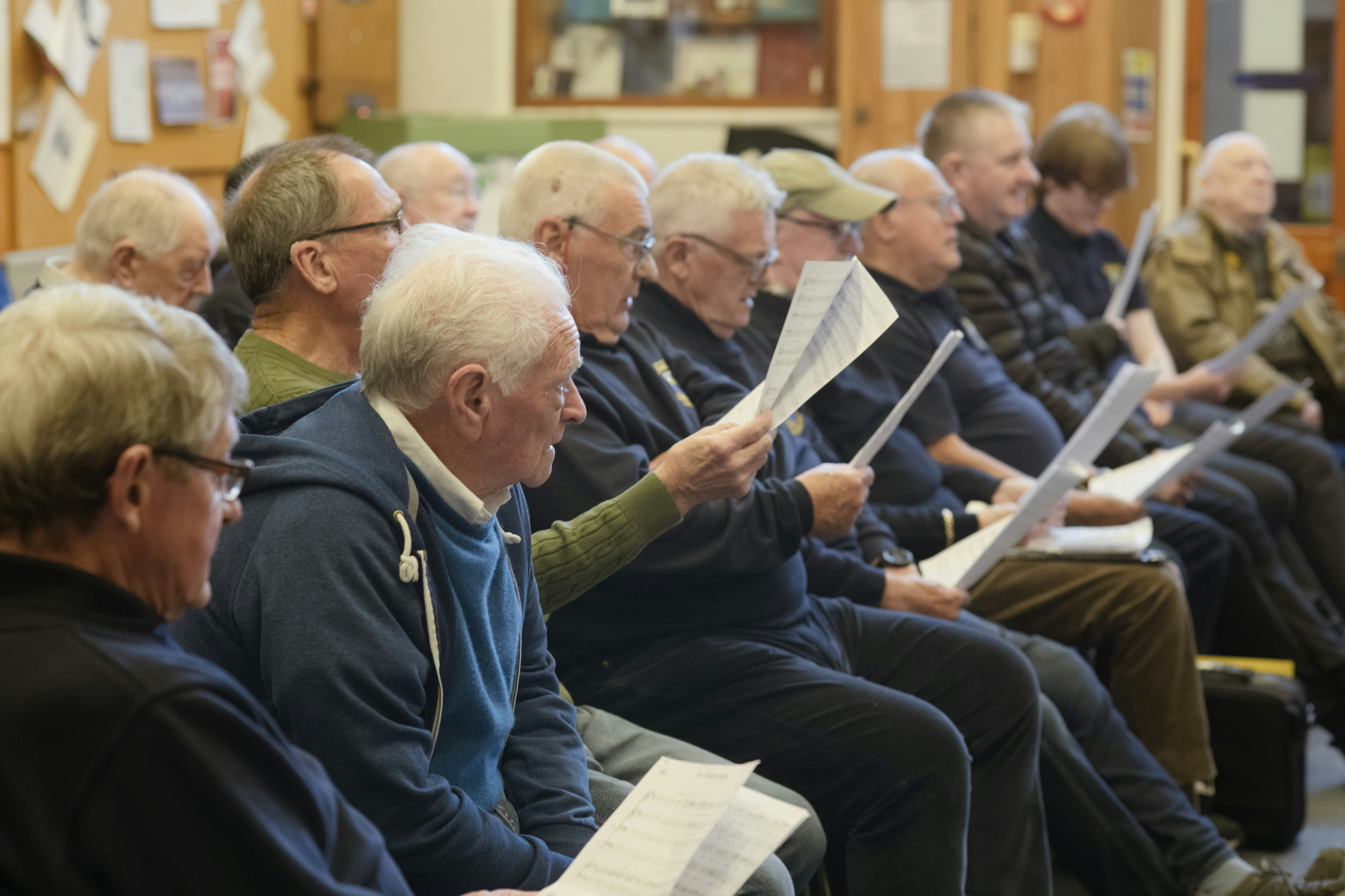 A group of elderly men seated in a hall, focused on sheet music and lyrics as they participate in a choir rehearsal.