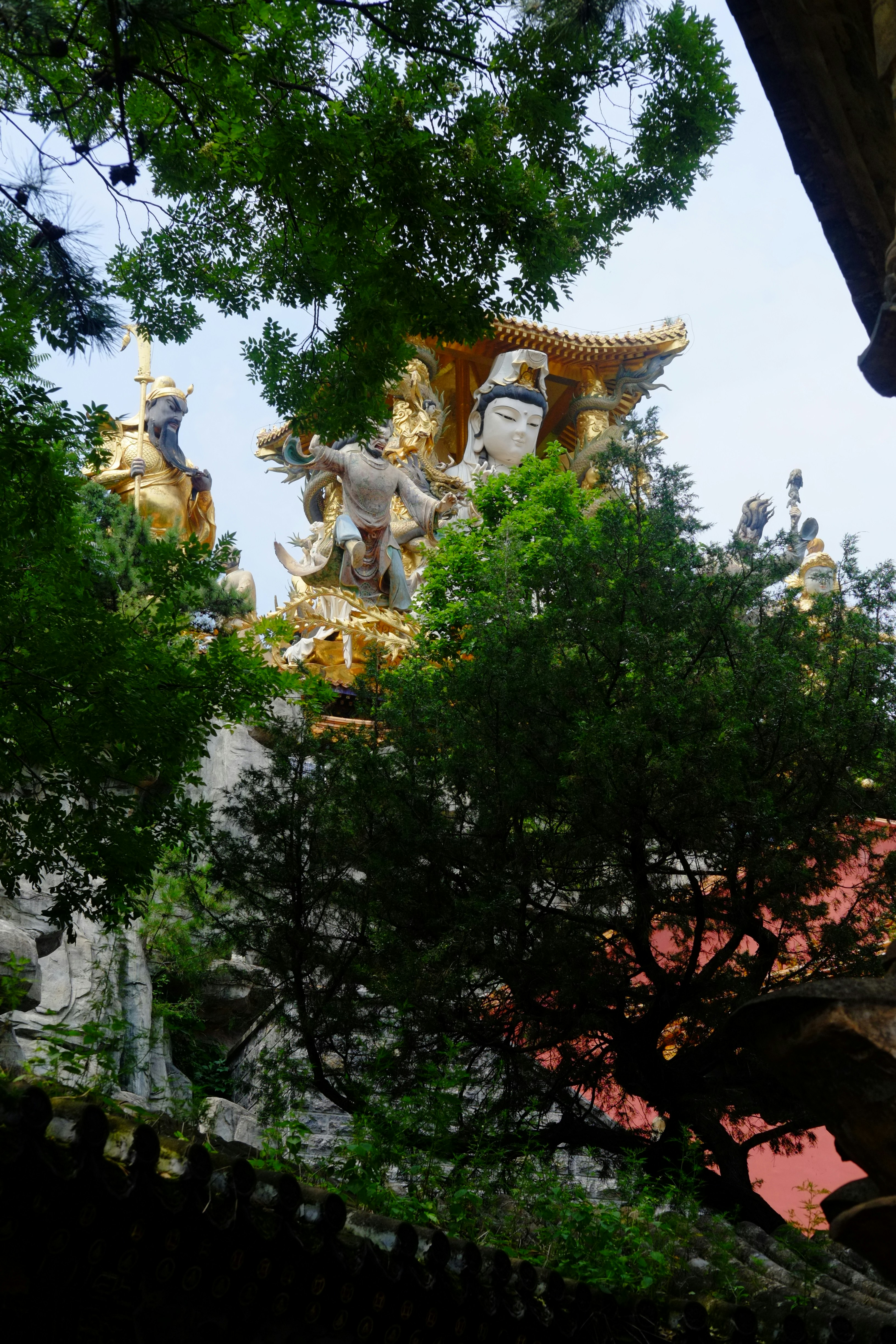 Stone and gold statues rise above a lush, leafy frame, peeking through trees toward a temple roof in the background.
