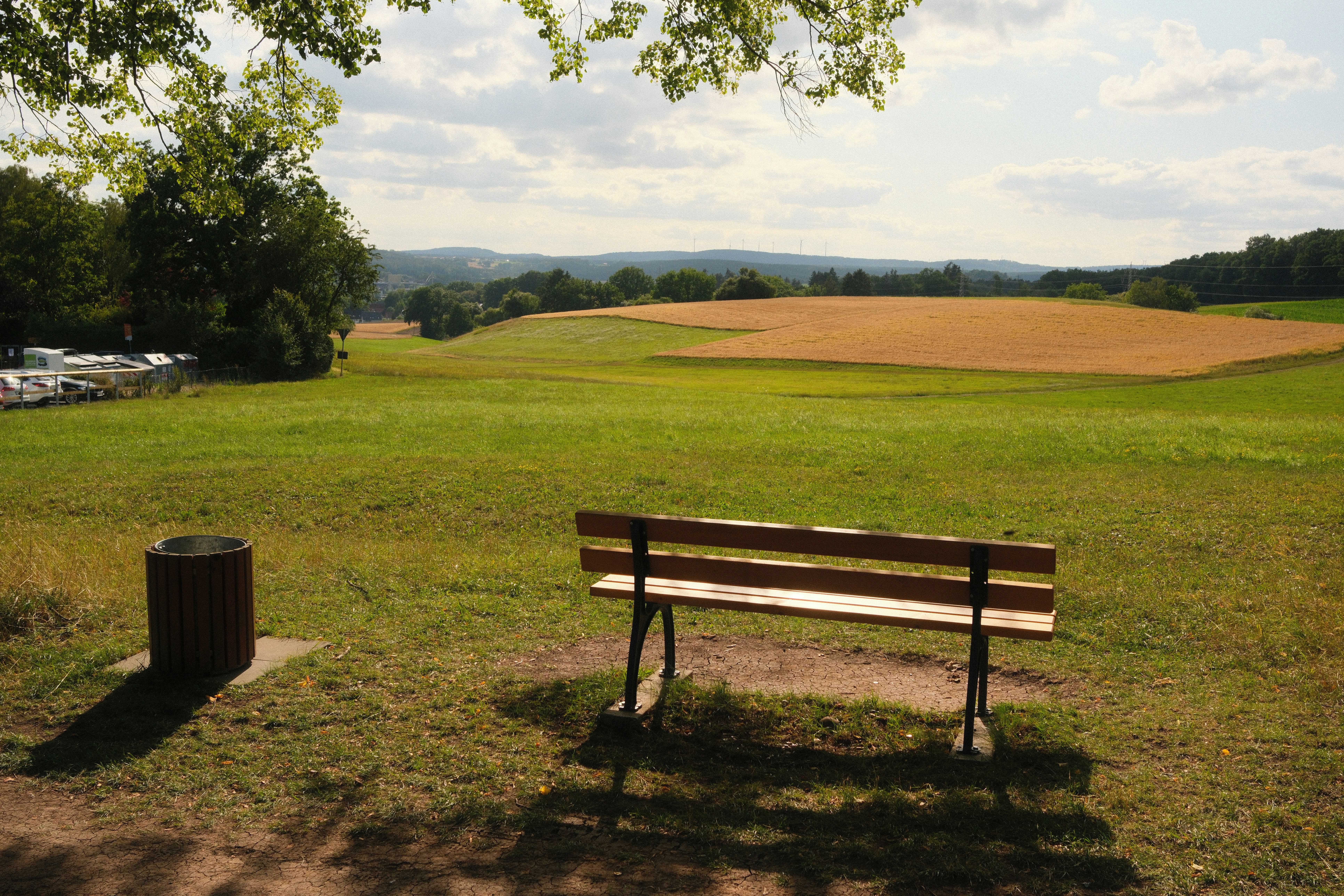 Sunlit park bench centered in a gentle landscape with rolling fields and distant hills; a trash can to the left anchors the scene as a quiet, everyday moment. (Photography evidence: outdoor plaza with bench and open sky.)