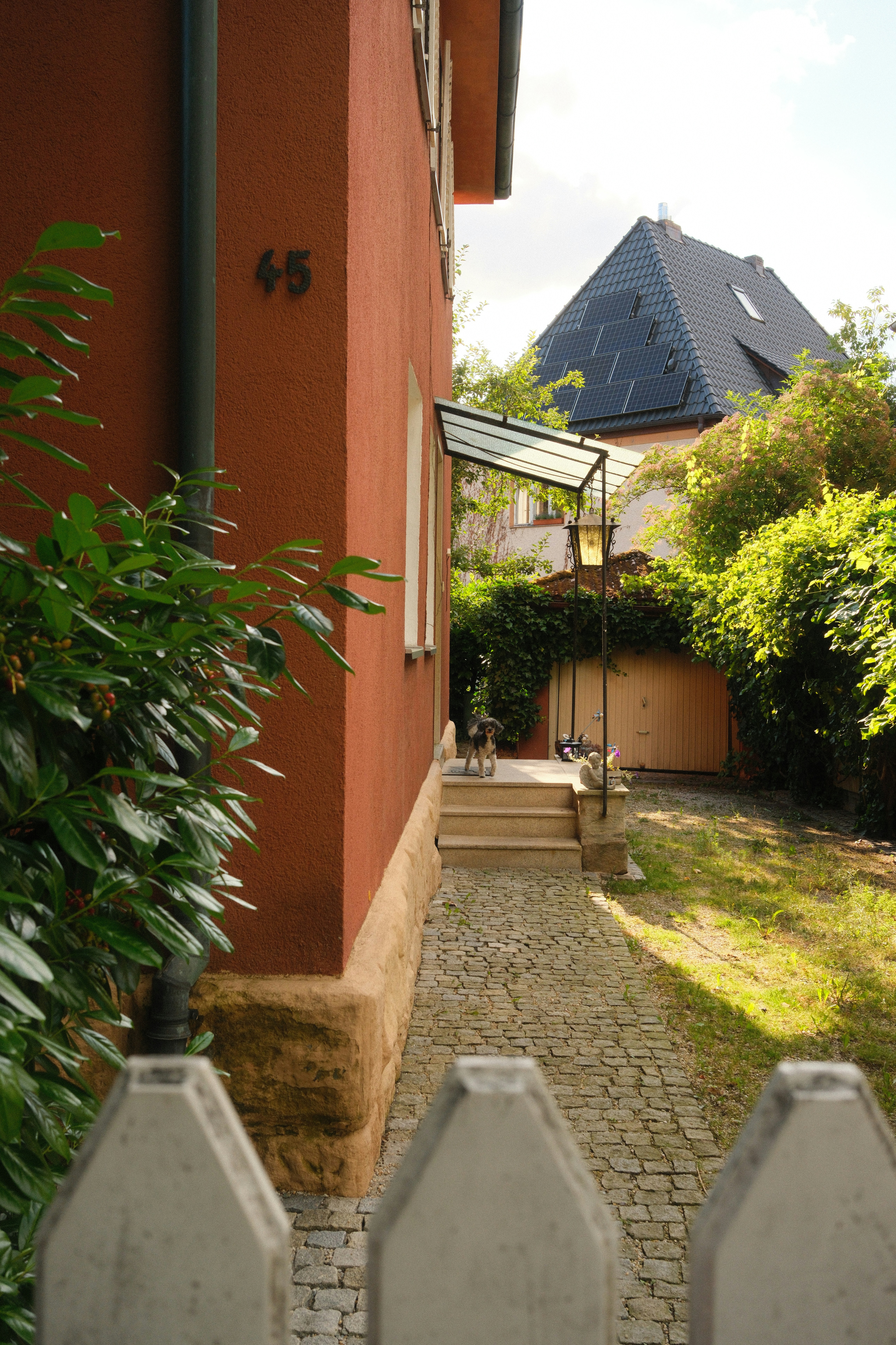 Paved path alongside a red house with a green garden.