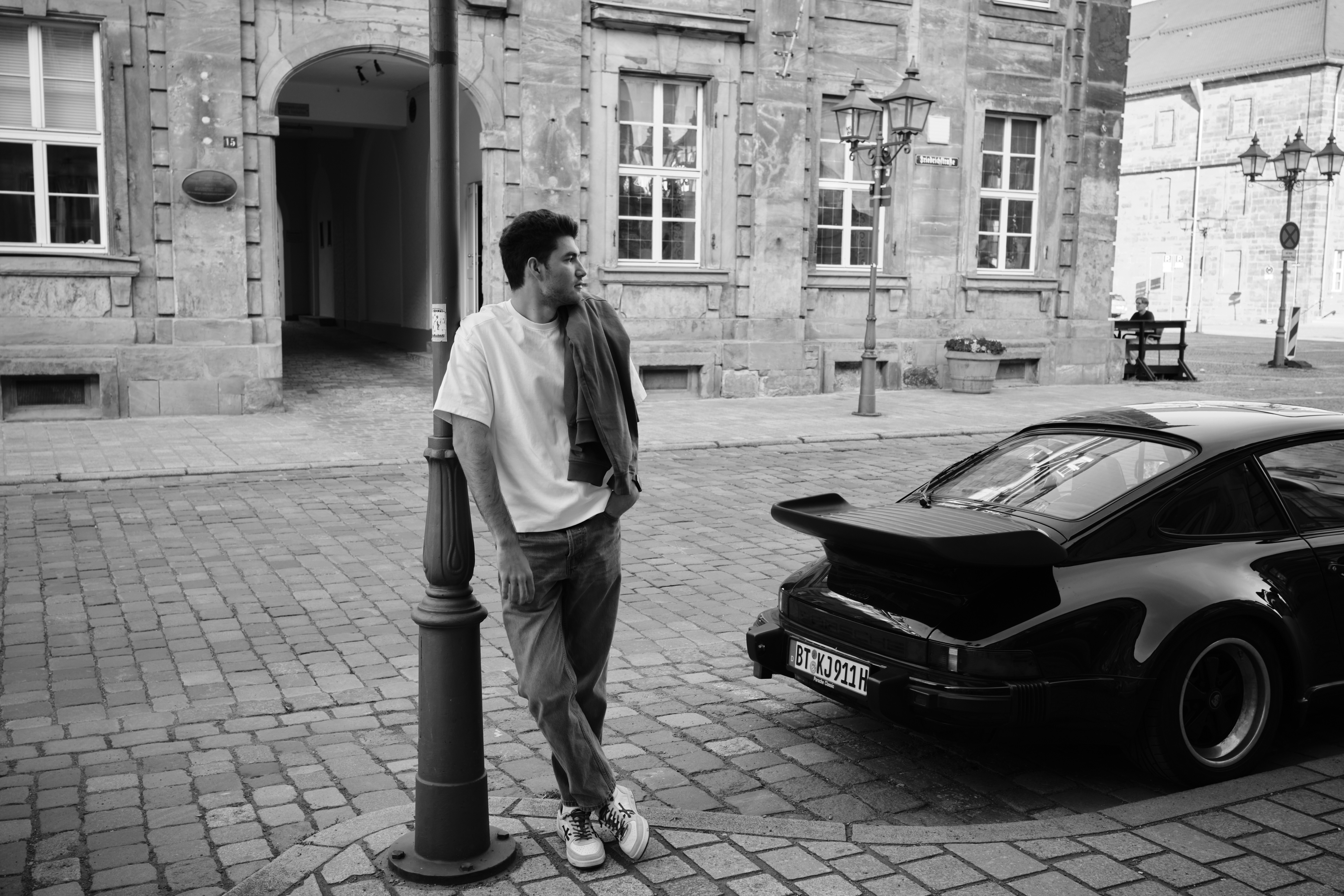 A man leans against a lamppost on a cobbled street beside a parked sports car, the grayscale scene capturing city texture and calm posture.