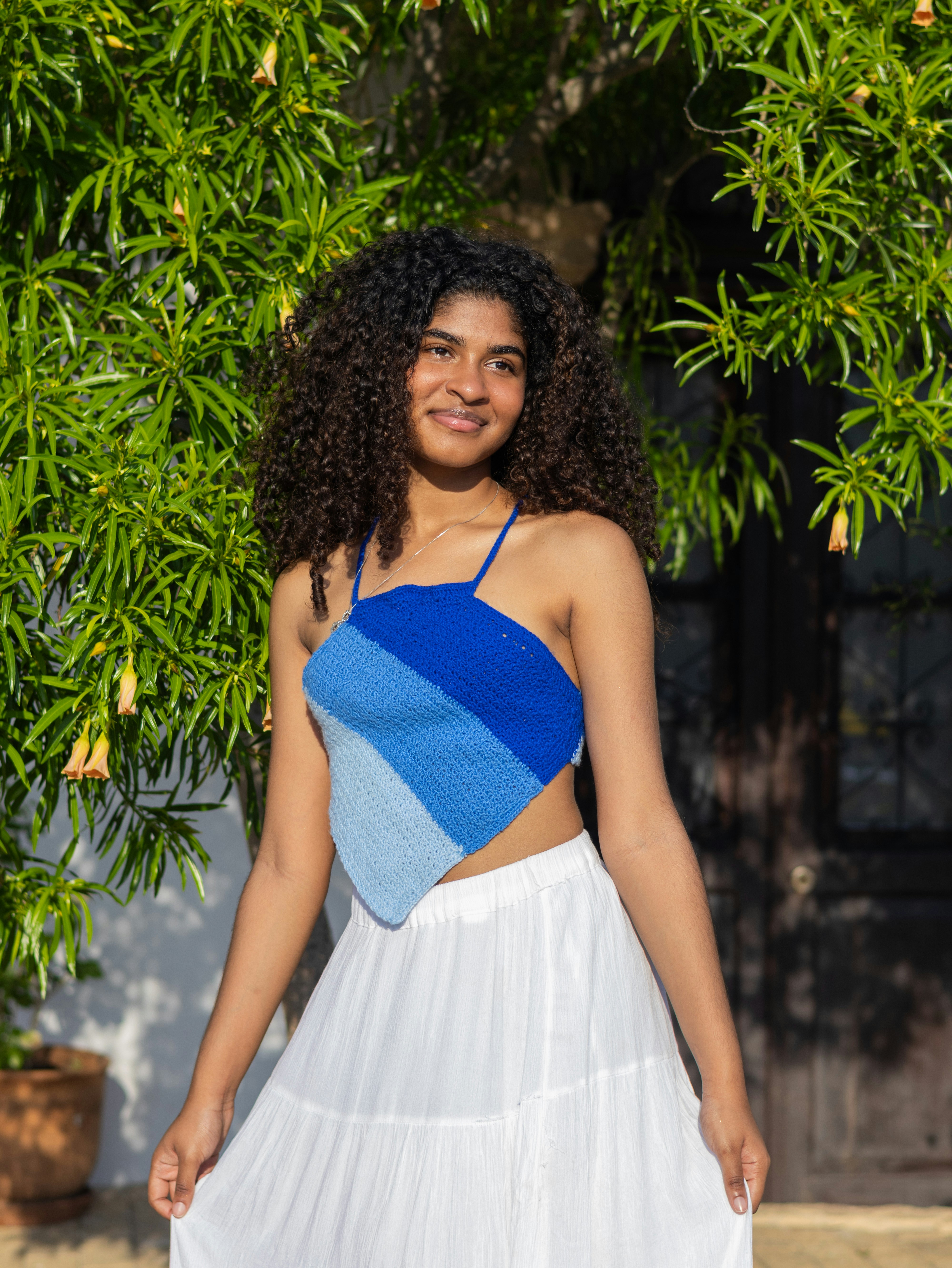 Sunlit portrait of a young woman in a blue crochet top and white skirt, standing under verdant hanging plants with a doorway behind. This is a lifestyle photograph capturing relaxed pose and natural light.