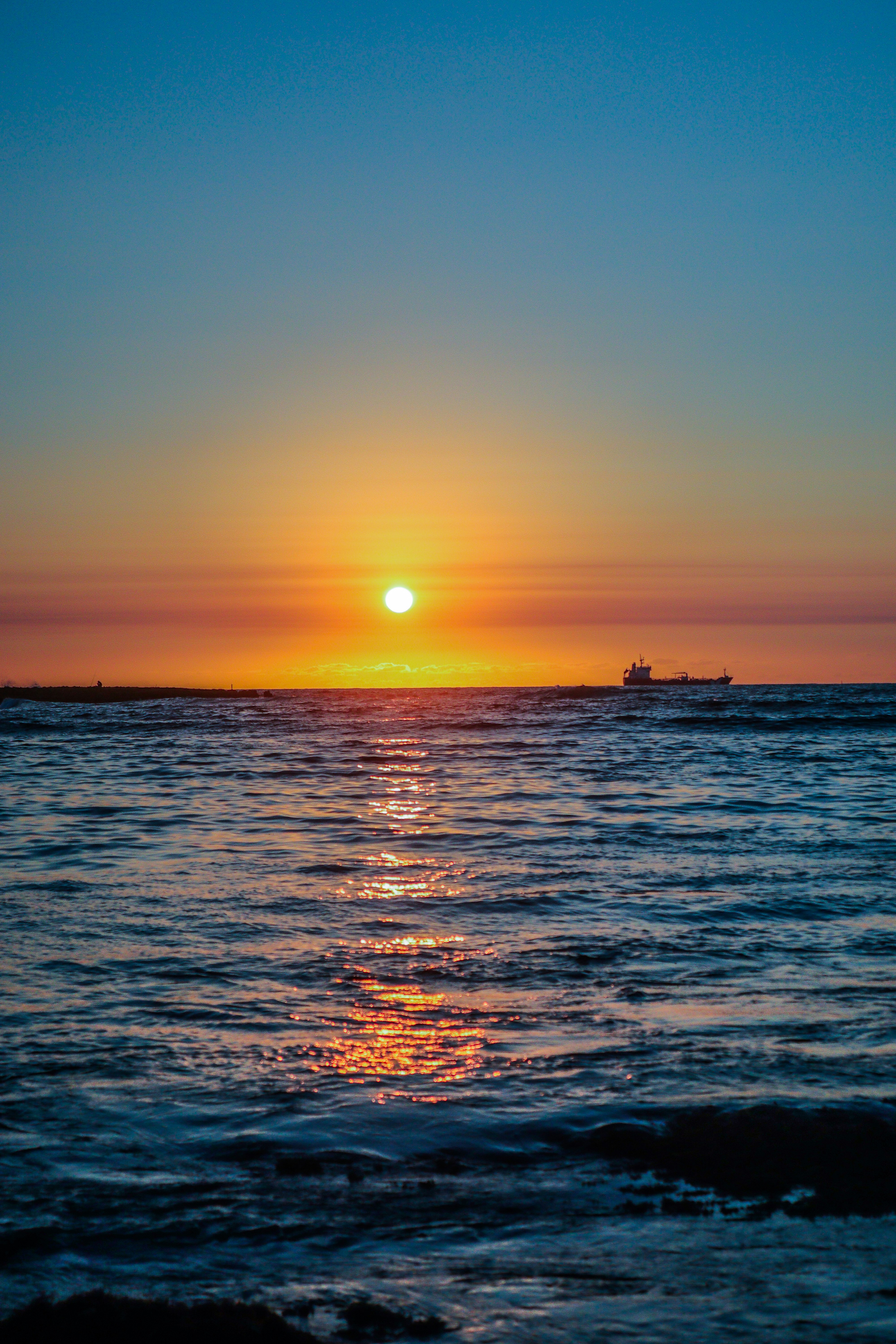 Vast ocean at sunset with a bright sun near the horizon and a distant ship on the right; water reflects warm tones, a calm, expansive seascape. This is a photographic capture, not a painting or graphic.