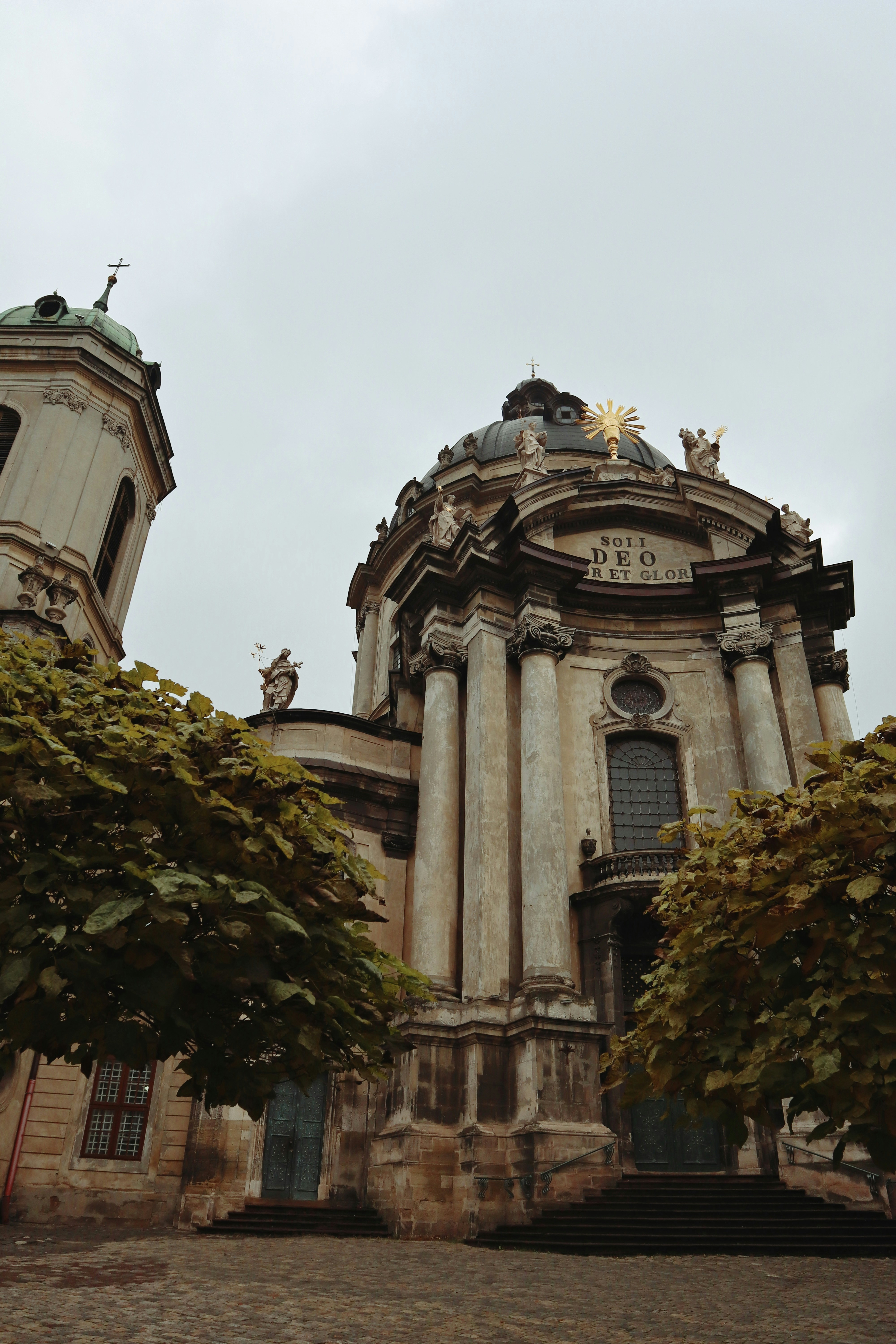 A magnificent vertical low-angle shot of the main facade of the historic Dominican Church (Church of the Holy Eucharist) in Lviv, Ukraine. The Baroque cathedral features impressive Corinthian columns, a grand dome adorned with a golden sunburst cross, and the Latin inscription "Soli Deo Honor et Gloria." Framed by green and yellow trees in the foreground against a moody gray sky, it captures the ancient architectural heritage of Eastern Europe.