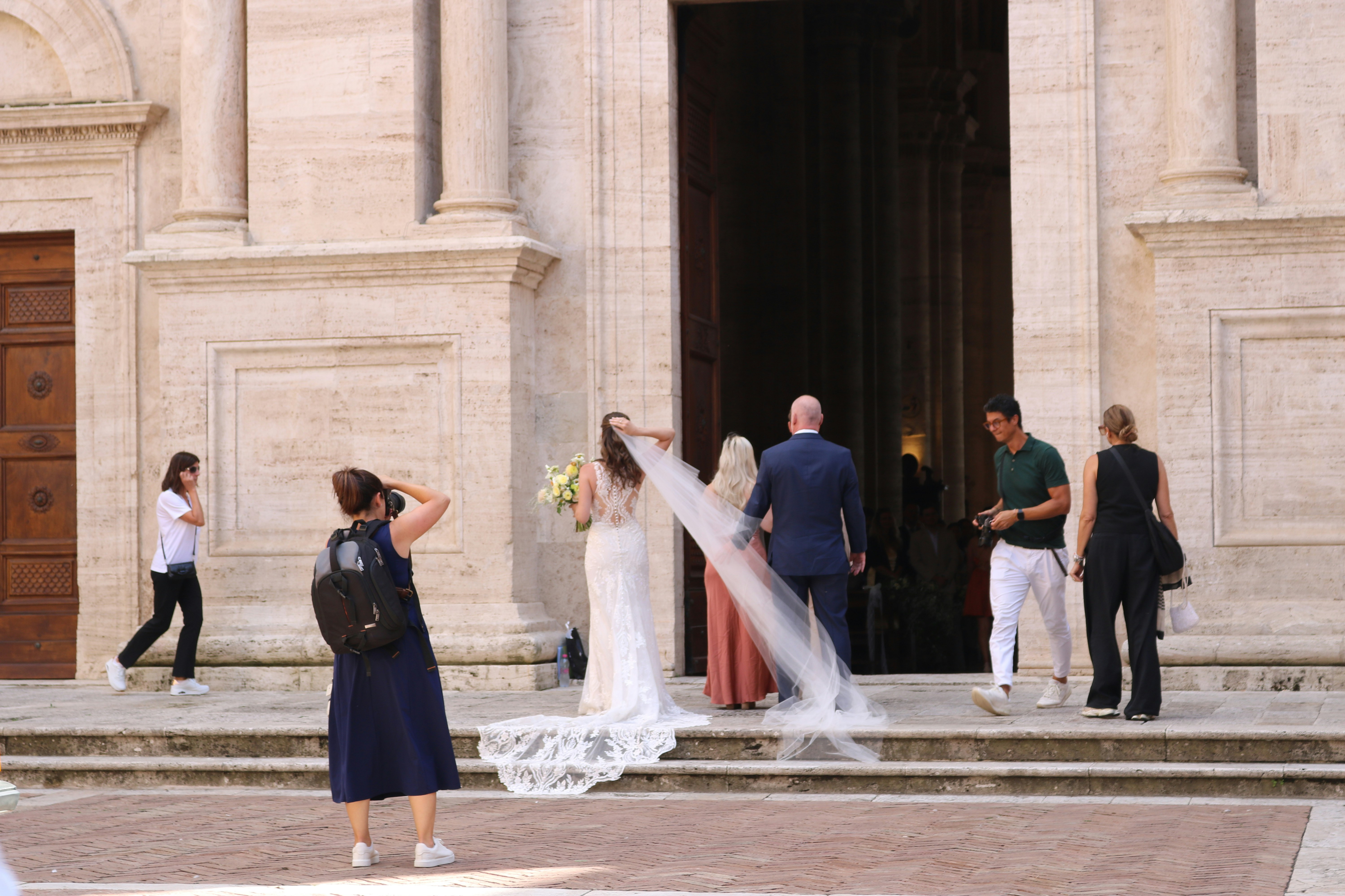 Bride and groom with veil trailing walk away from a grand entrance, surrounded by guests on the steps of a stone building.